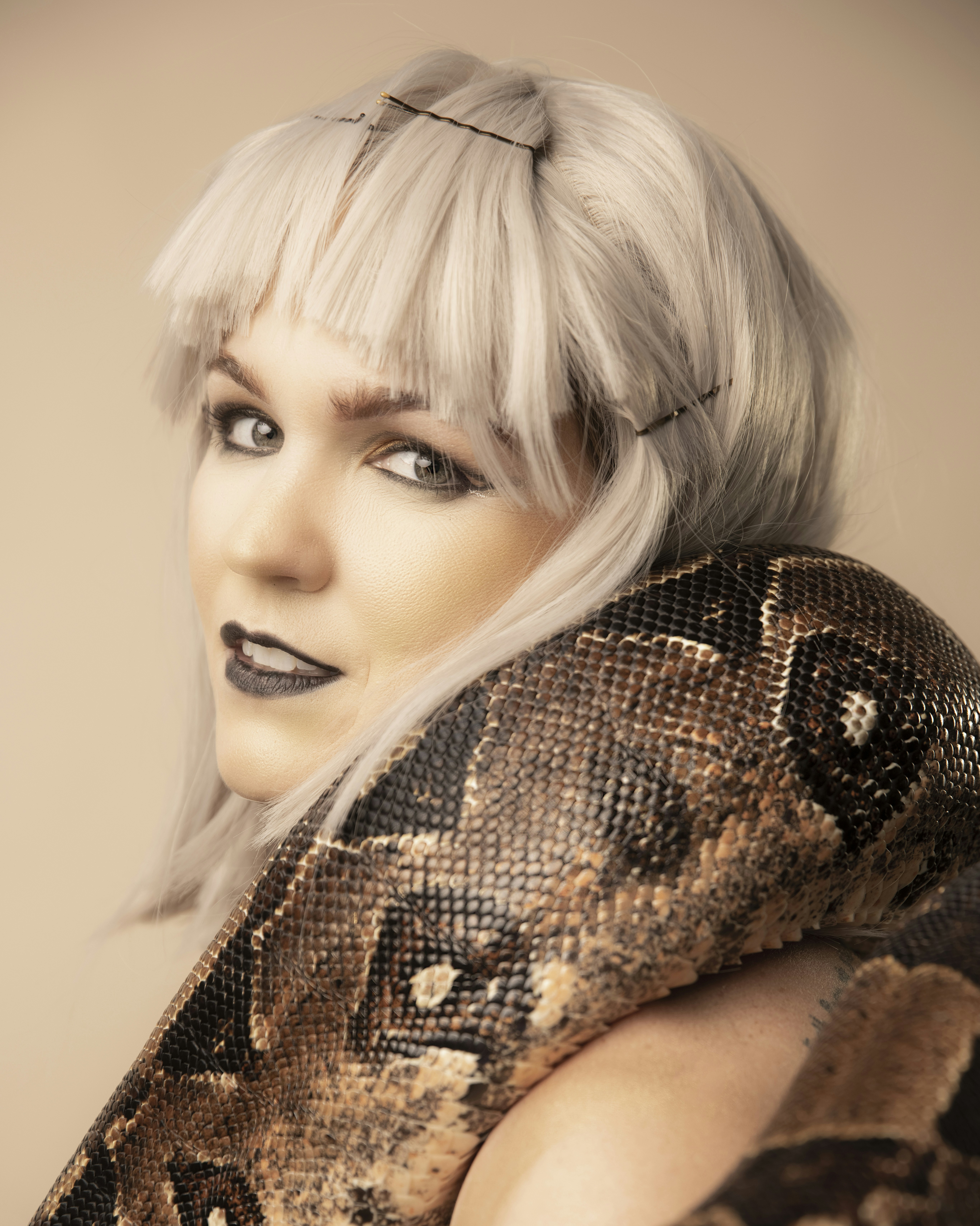 A model with silver hair and dark makeup poses with a large snake draped around her shoulders, creating a striking contrast against a soft background.