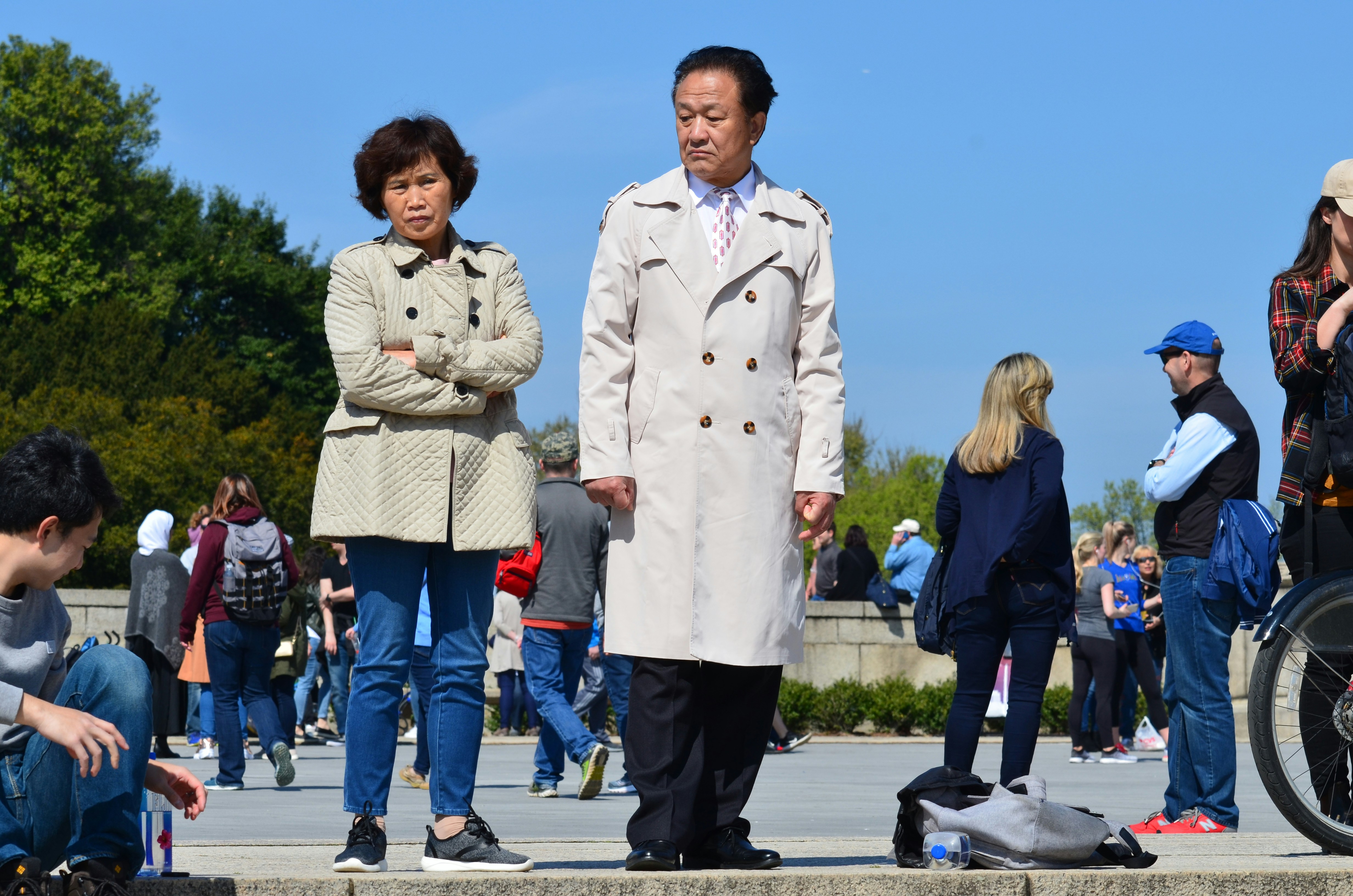 Two individuals stand thoughtfully in a bustling public space, surrounded by a diverse group of people enjoying a sunny day.