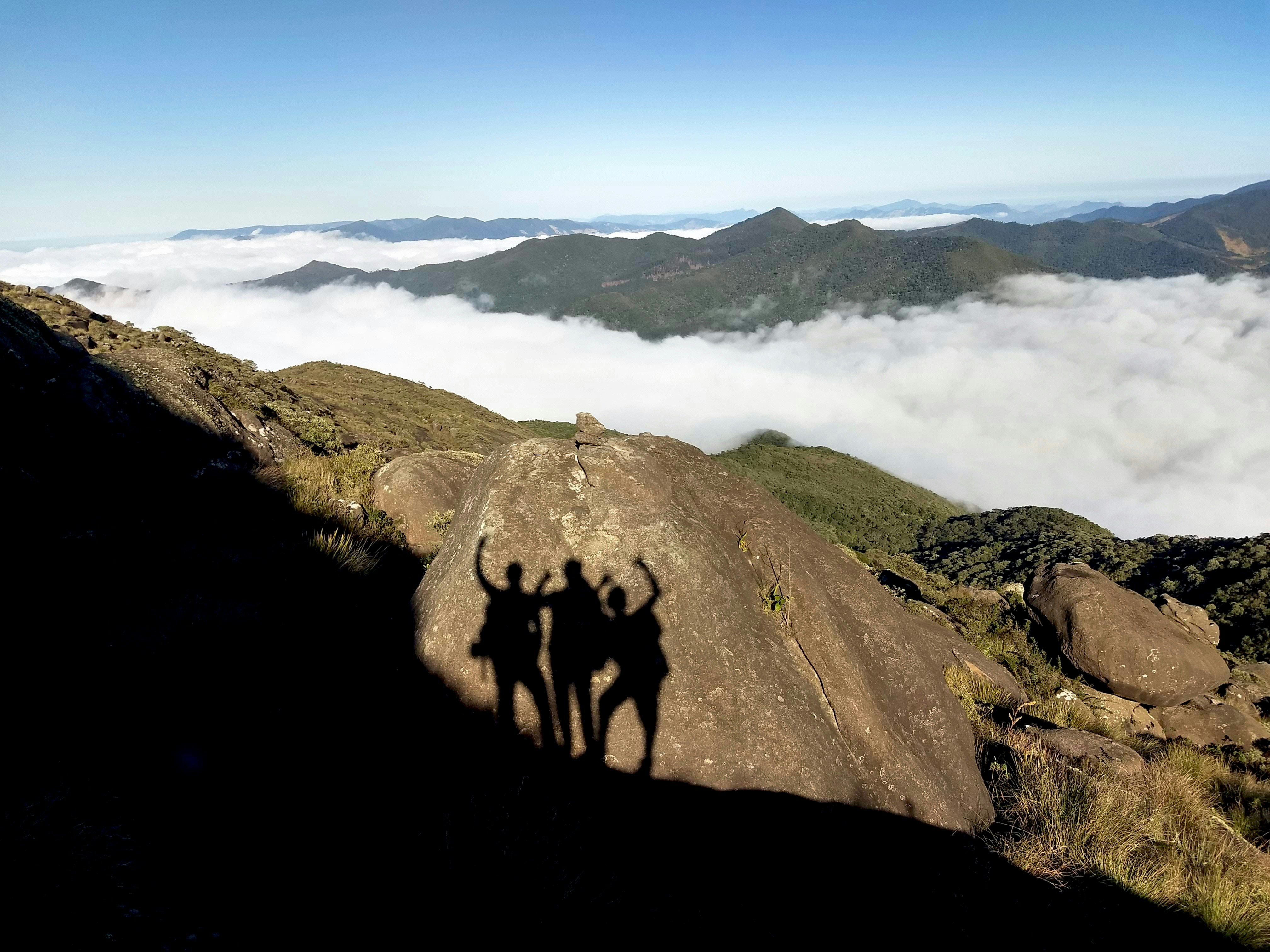 Three hikers' silhouettes stand on a rocky ridge above a sea of clouds at dawn.