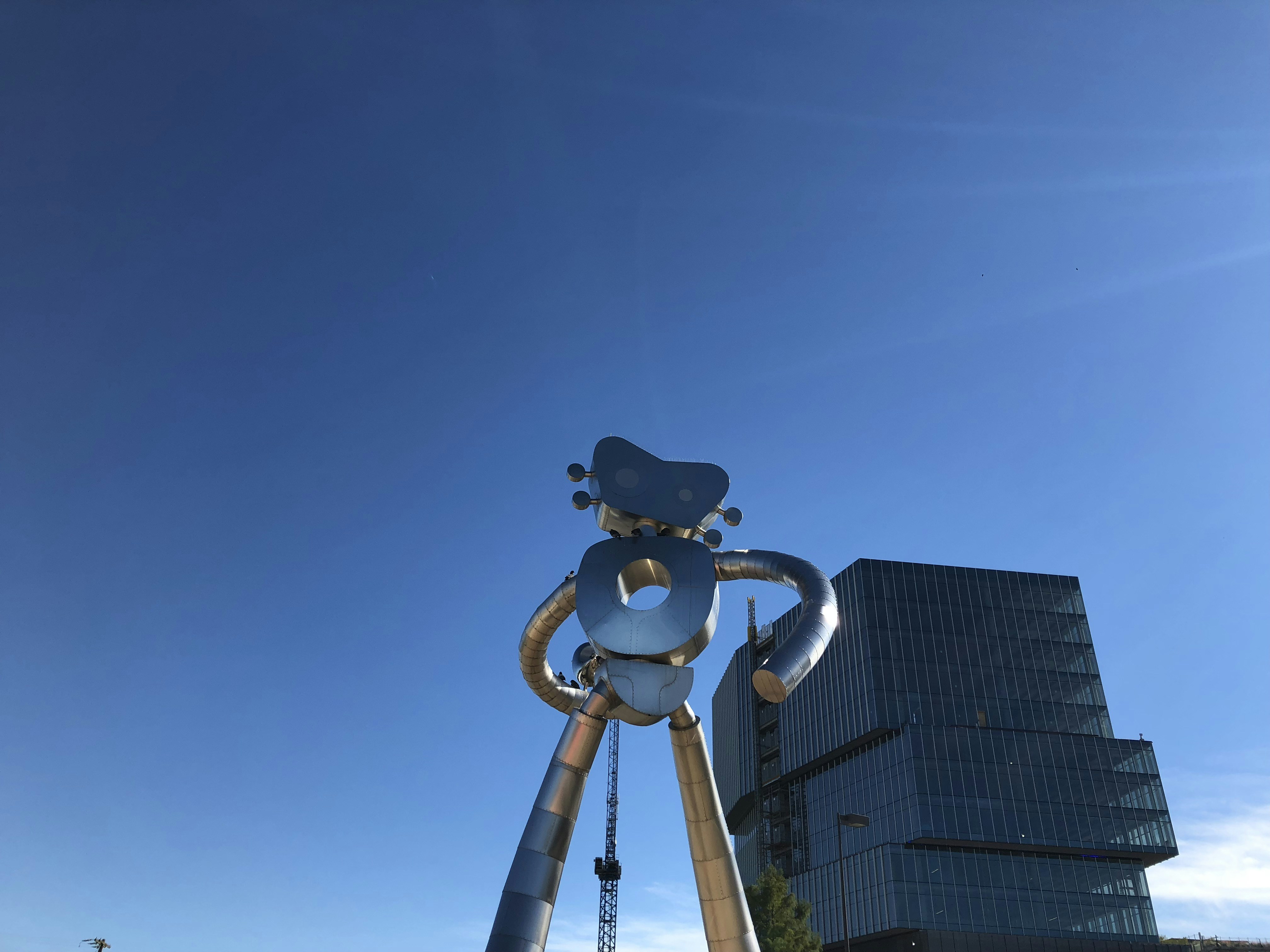 Abstract metal sculpture with circular elements against a bright blue sky, adjacent to a modern glass building.