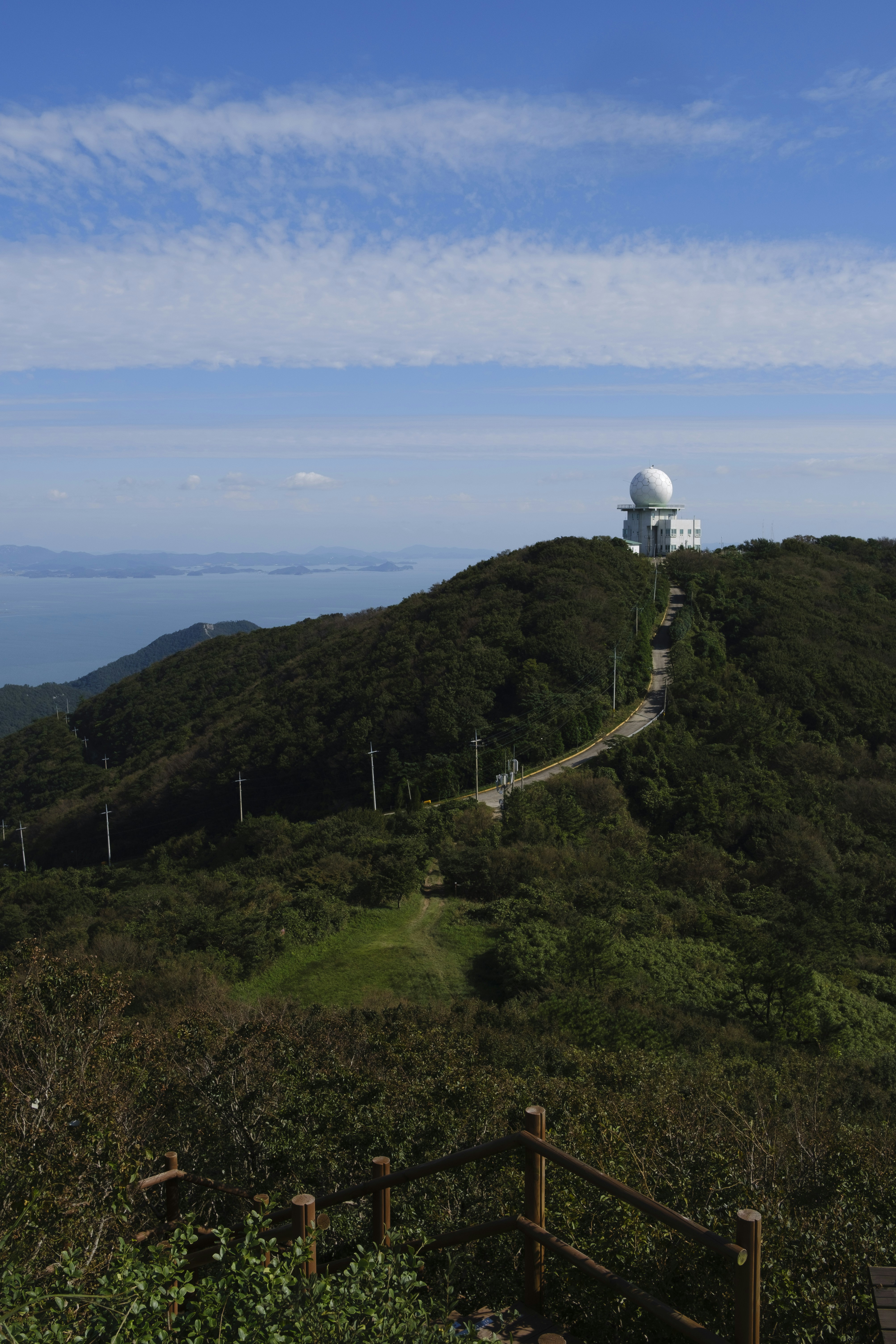 A radar tower perched atop a lush green mountain, overlooking a tranquil sea under a blue sky with scattered clouds.