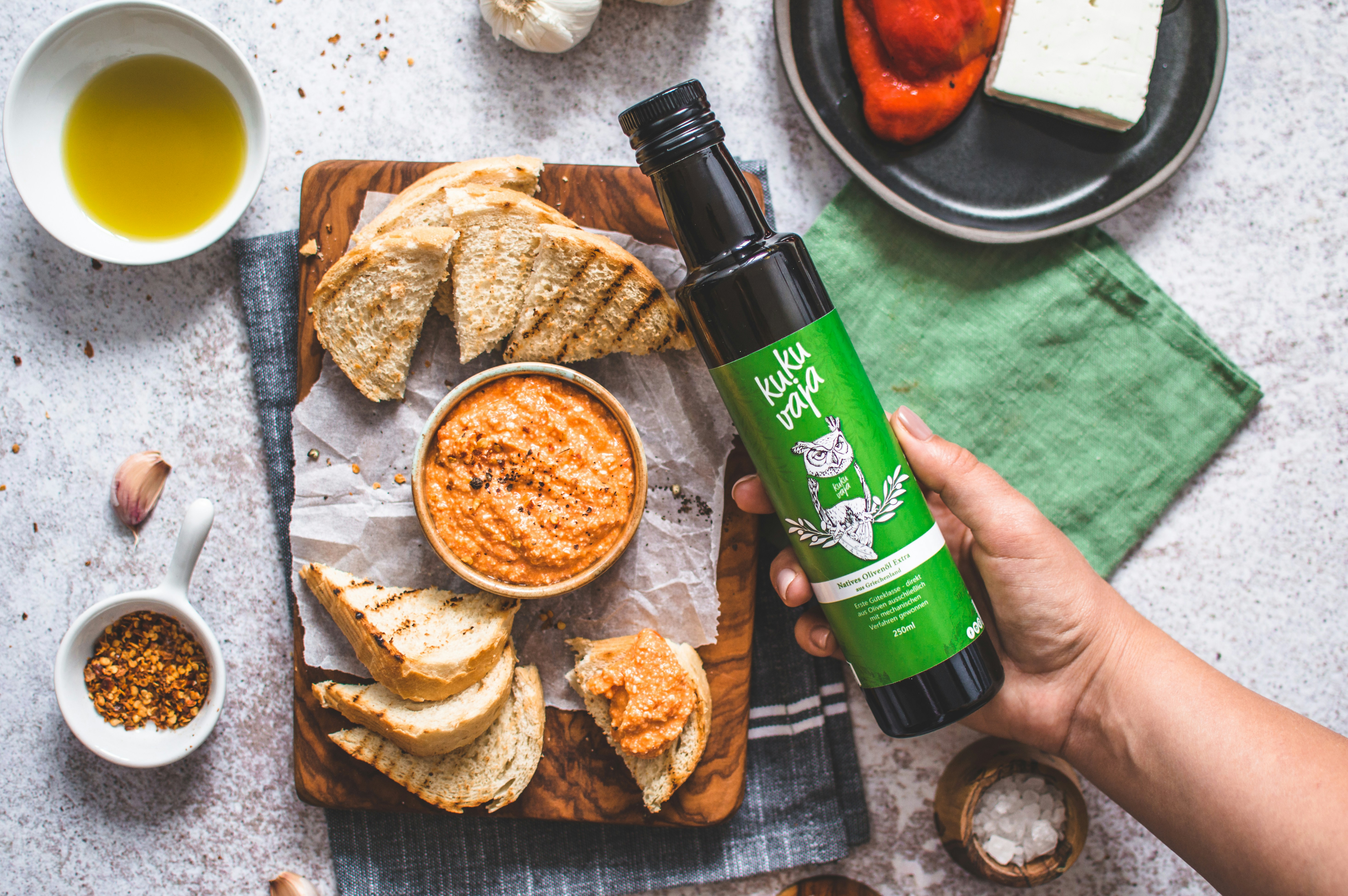 Hand holding a green bottle of olive oil next to sliced bread and a bowl of spread on a wooden board.