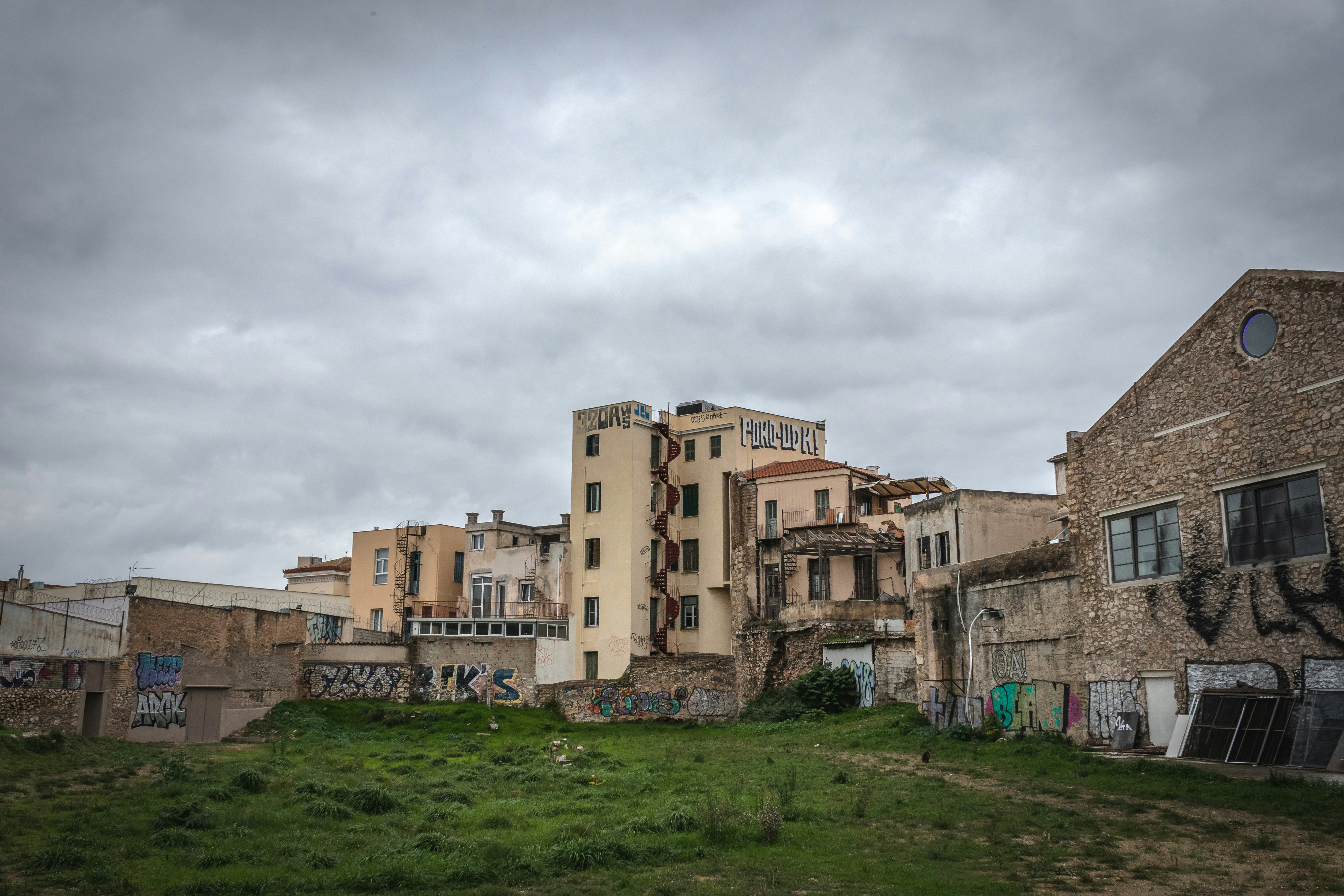 Abandoned buildings adorned with graffiti stand amidst overgrown grass under a cloudy sky, reflecting the passage of time and neglect.