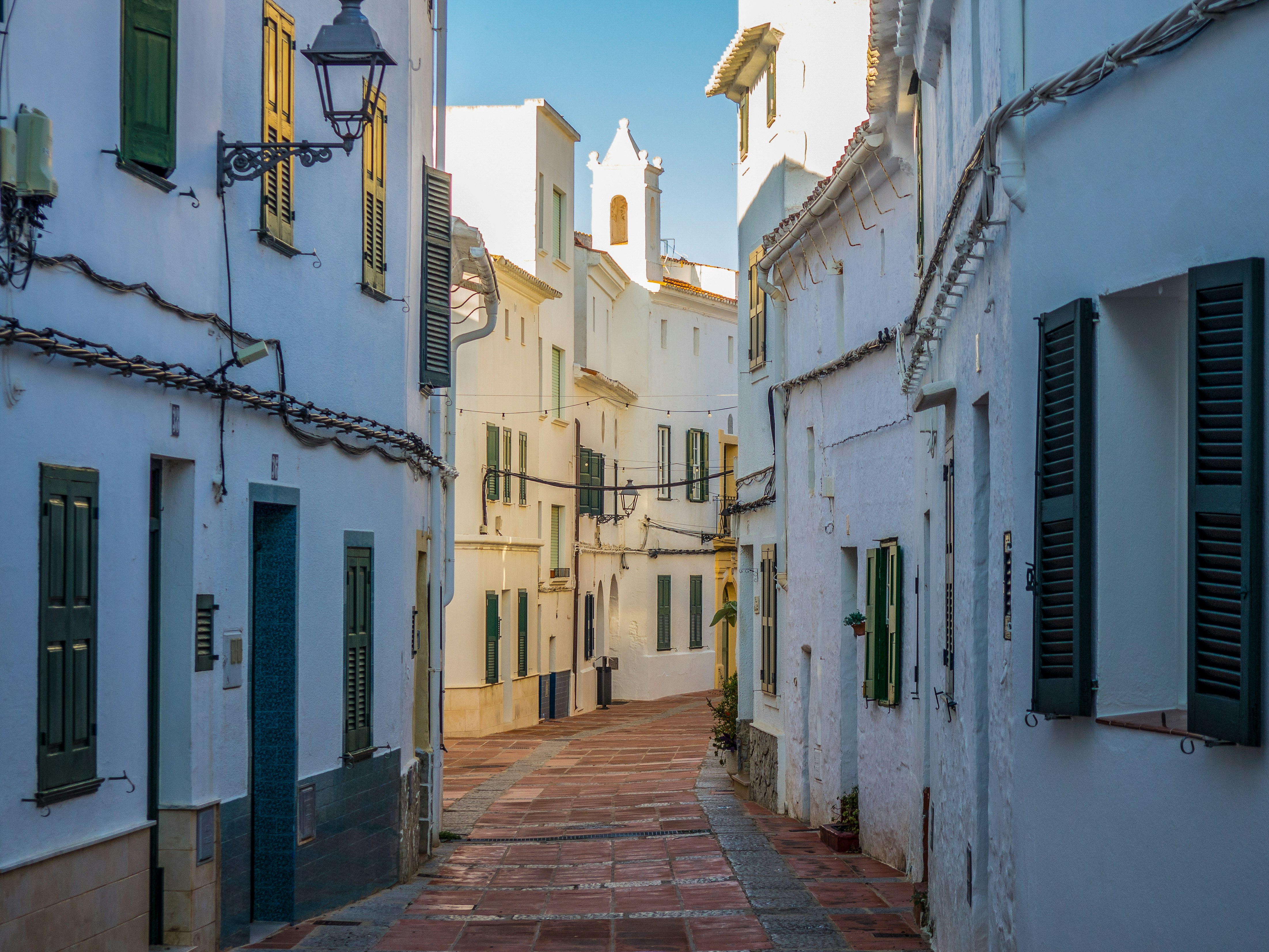 Narrow cobblestone street lined with whitewashed buildings and green shutters under a clear blue sky.