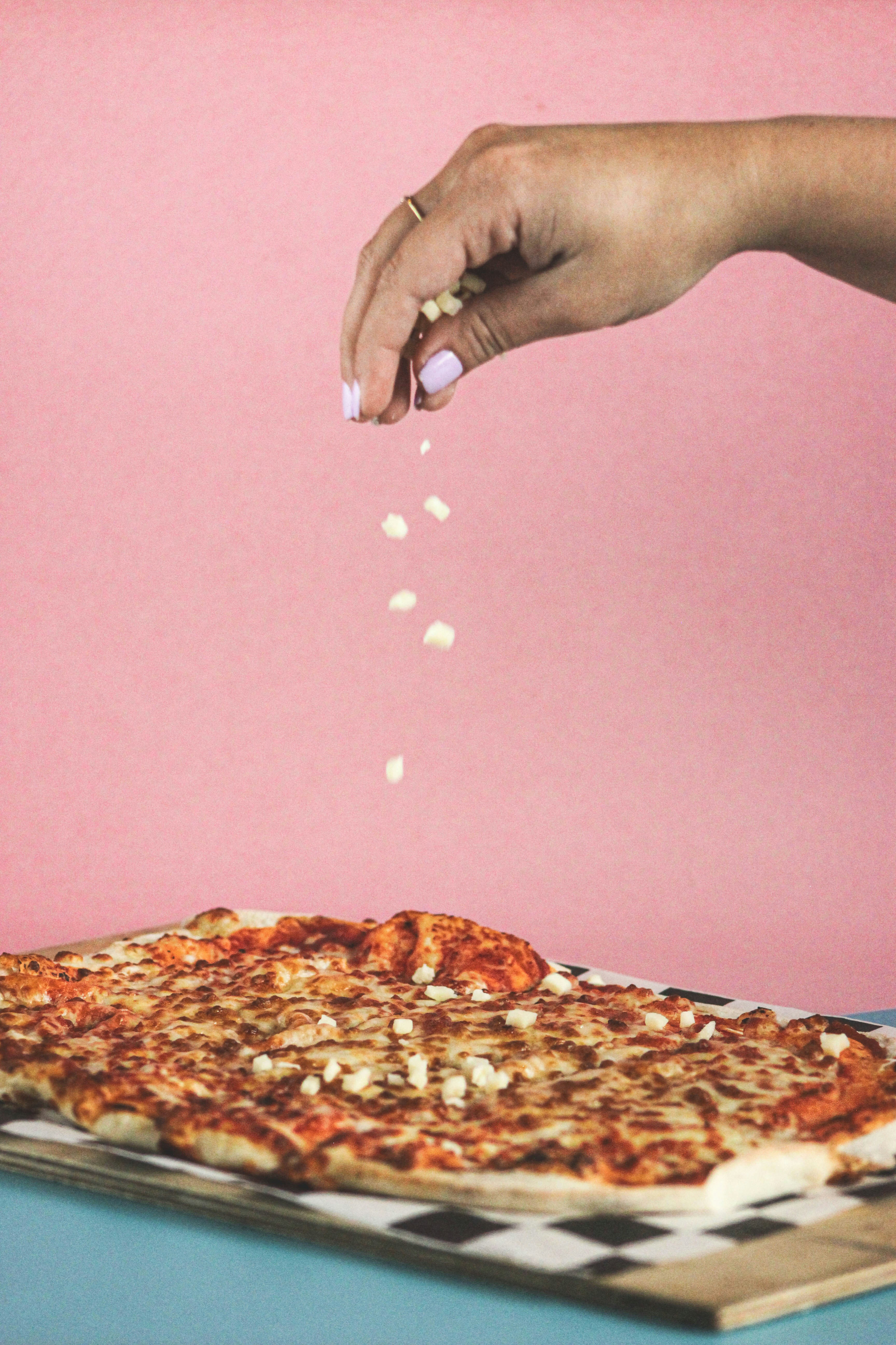Hand sprinkling toppings over a freshly baked pizza on a checkered board with a pink backdrop.