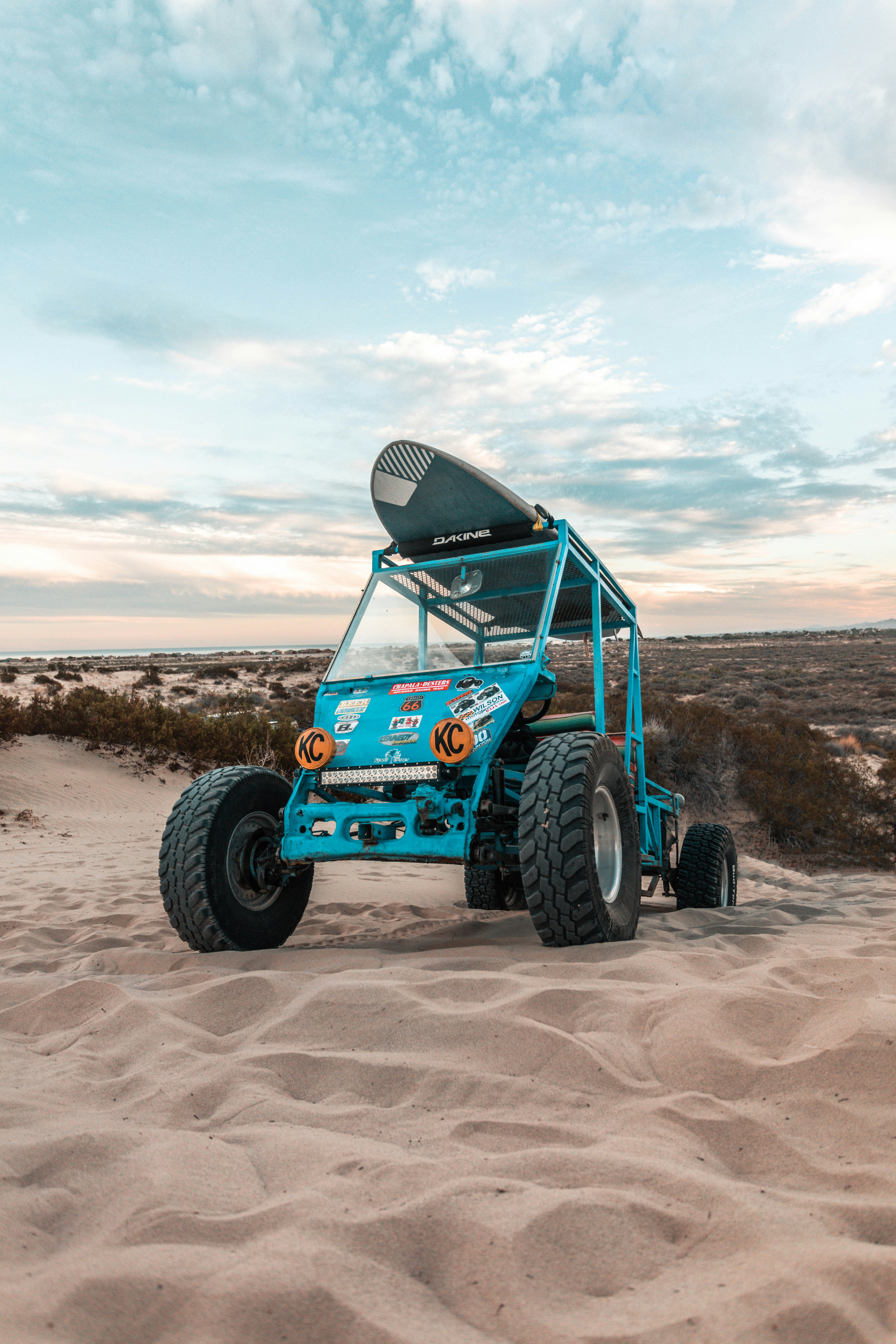 A vibrant blue beach buggy parked on sandy dunes, topped with a surfboard, set against a dramatic sky. The scene evokes a sense of adventure and leisure.