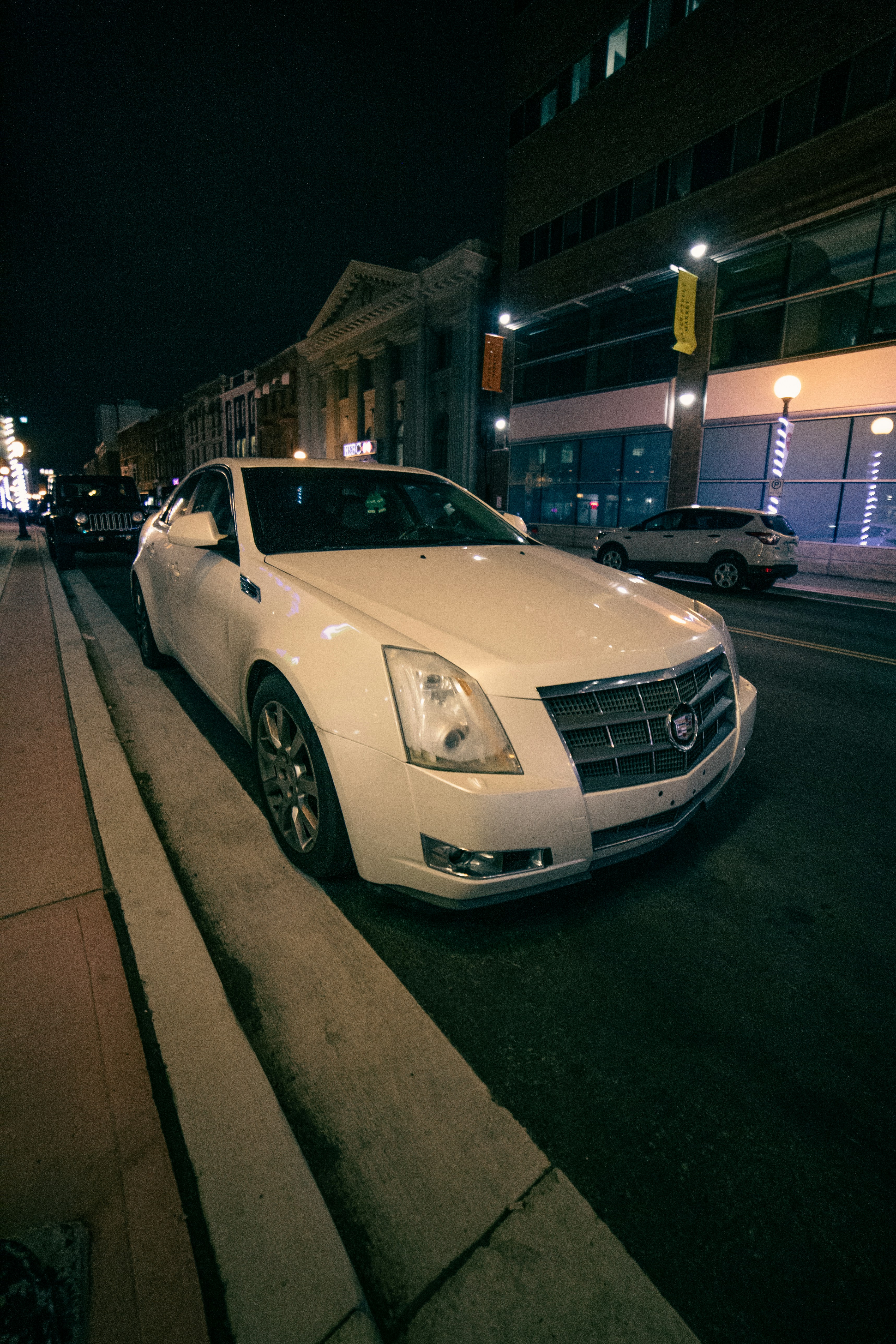 White Cadillac parked along a city street at night, illuminated by nearby lights. The urban backdrop adds depth to the scene.
