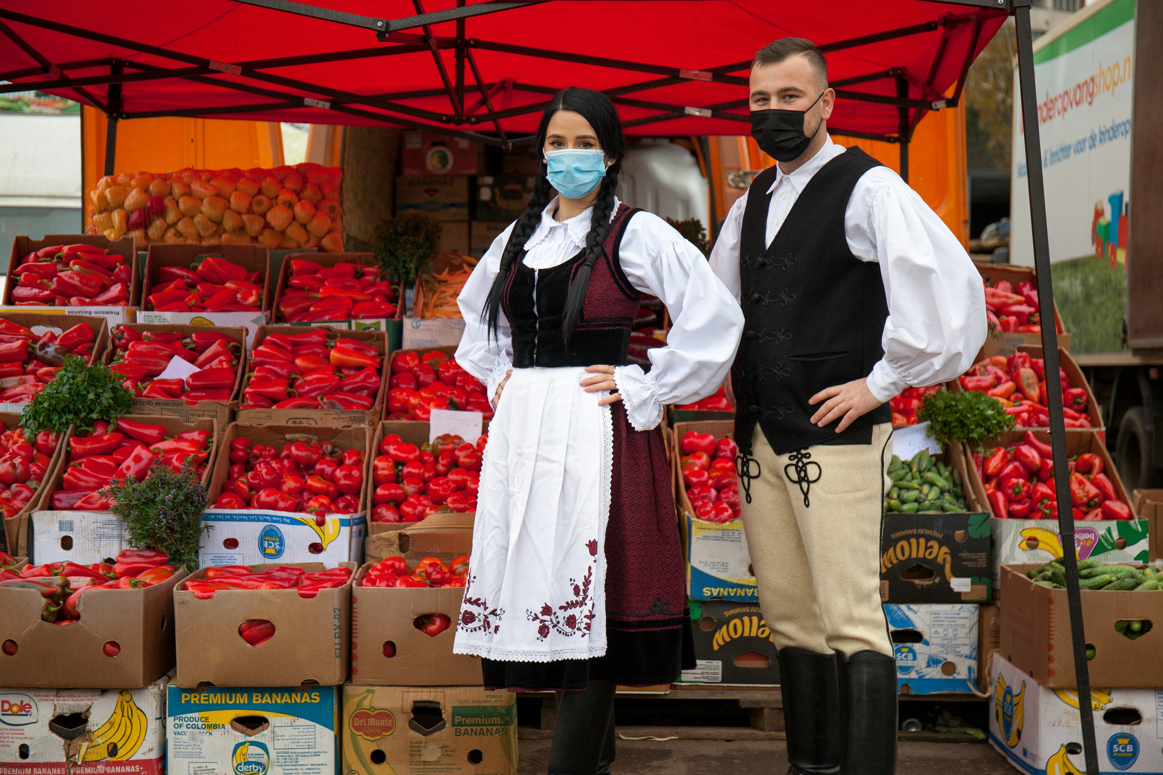 Two individuals in traditional attire stand before a vibrant display of produce at an outdoor market.