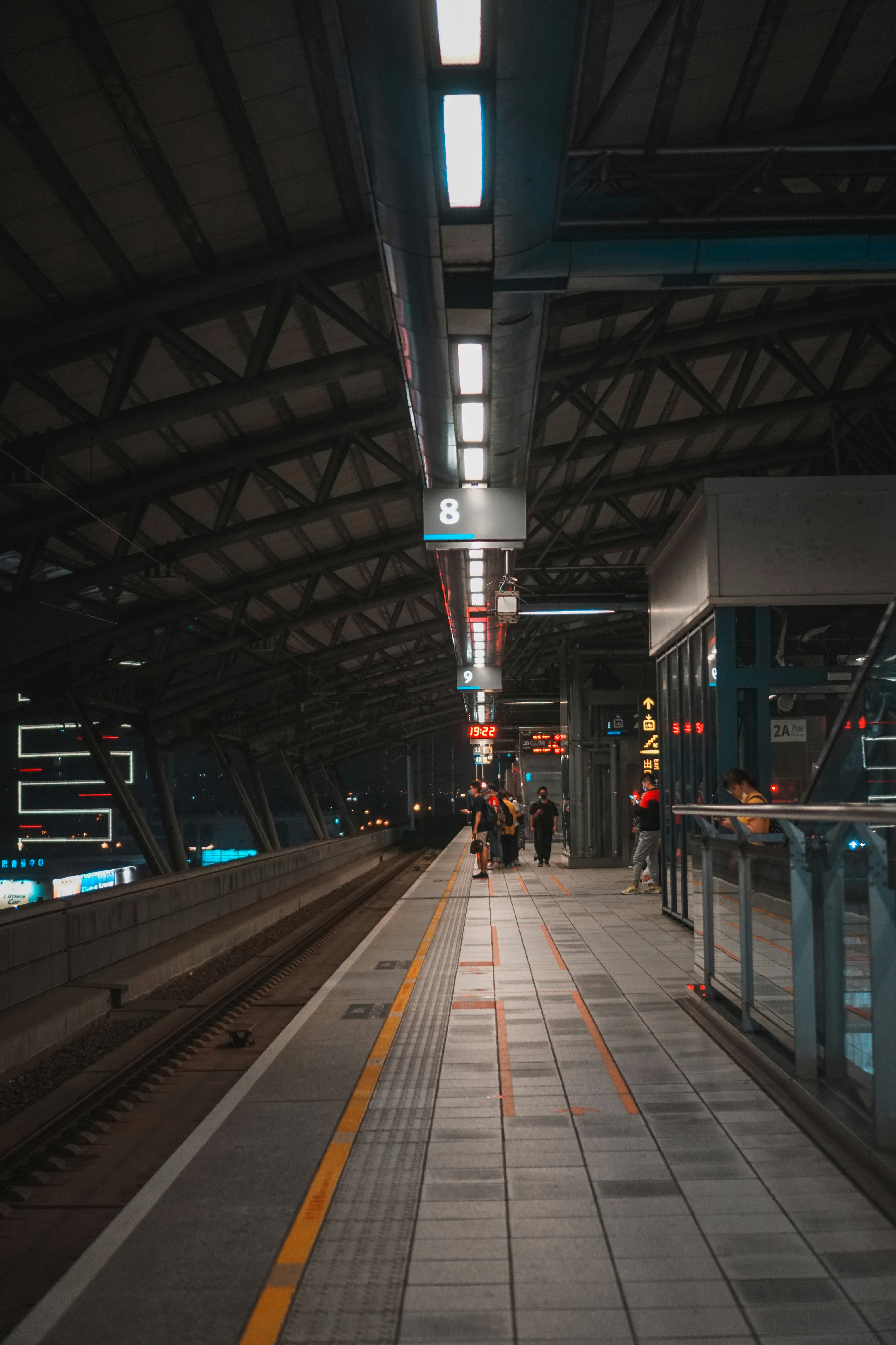 A modern train platform illuminated by overhead lights, showcasing waiting passengers and digital displays. The architectural lines create a dynamic perspective.
