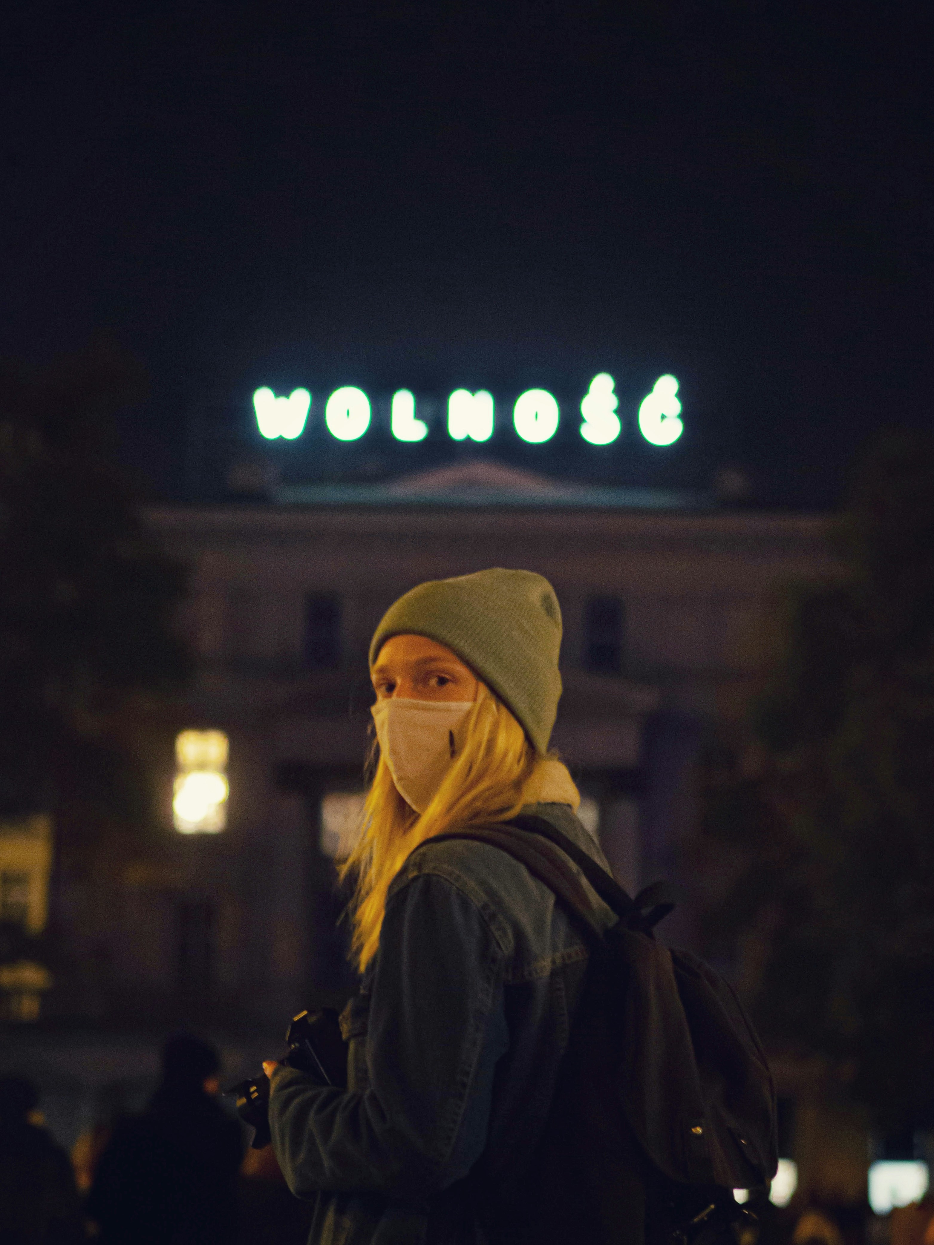 Person in a beanie and mask stands in front of a building with a glowing sign reading 'Wolność' at night.