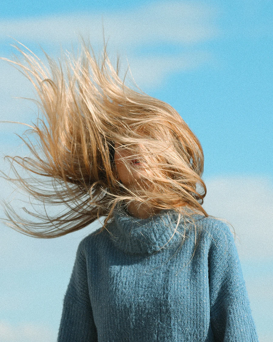 A person in a textured blue turtleneck with long hair whipped across their face against a bright sky-blue background. The hair motion and soft light create a breezy, editorial feel.