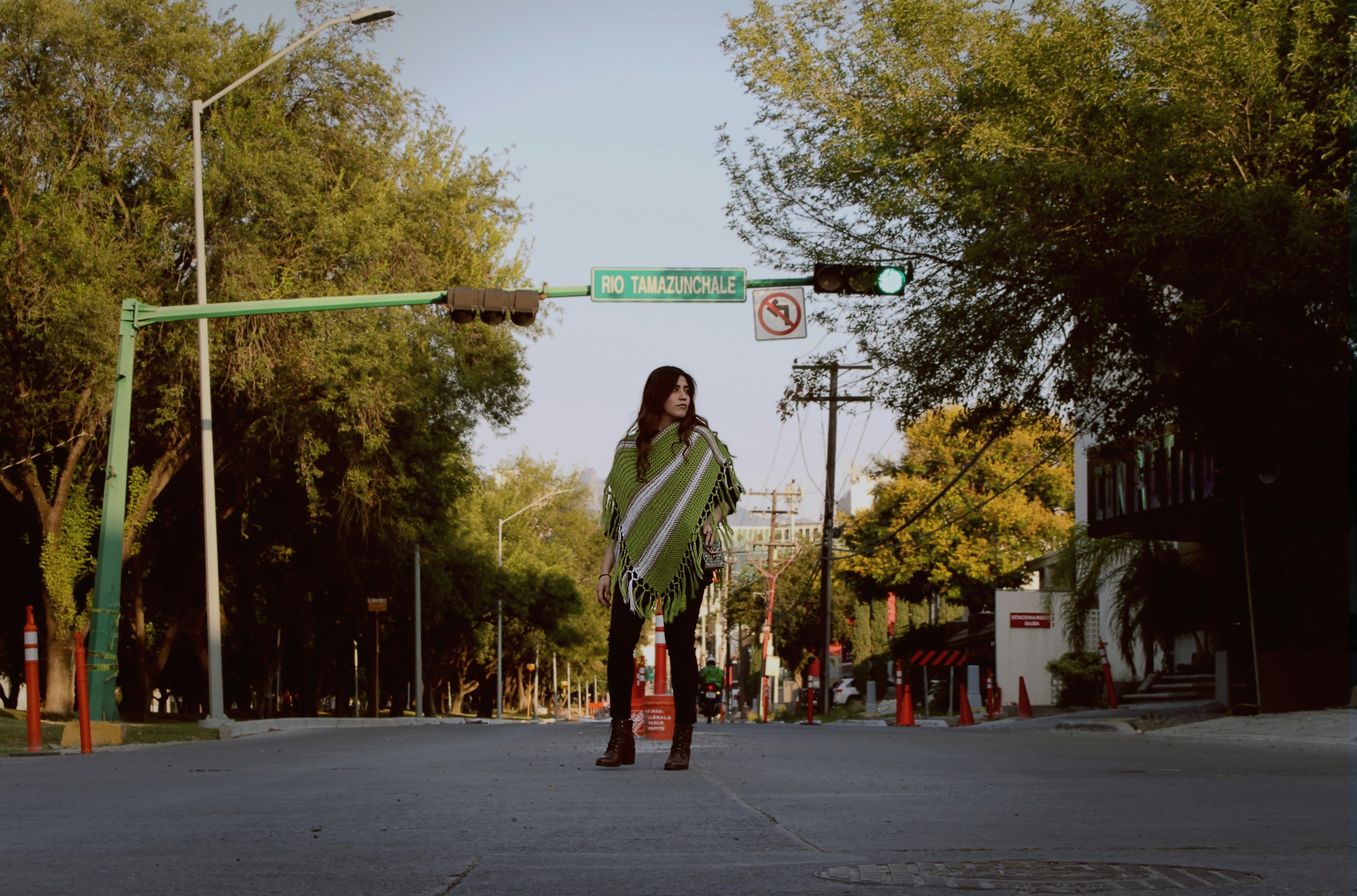 Person in a green poncho standing on a quiet street under a traffic light with trees lining the road.