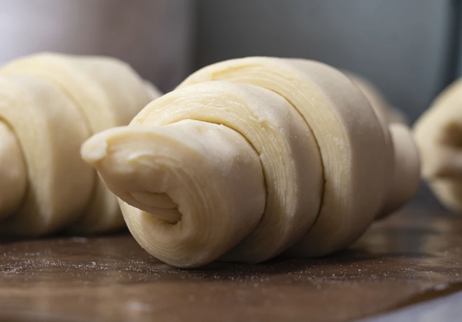 Close-up of raw croissant dough rolls resting on a floured wooden surface, with one centered in sharp focus and others blurred behind it.