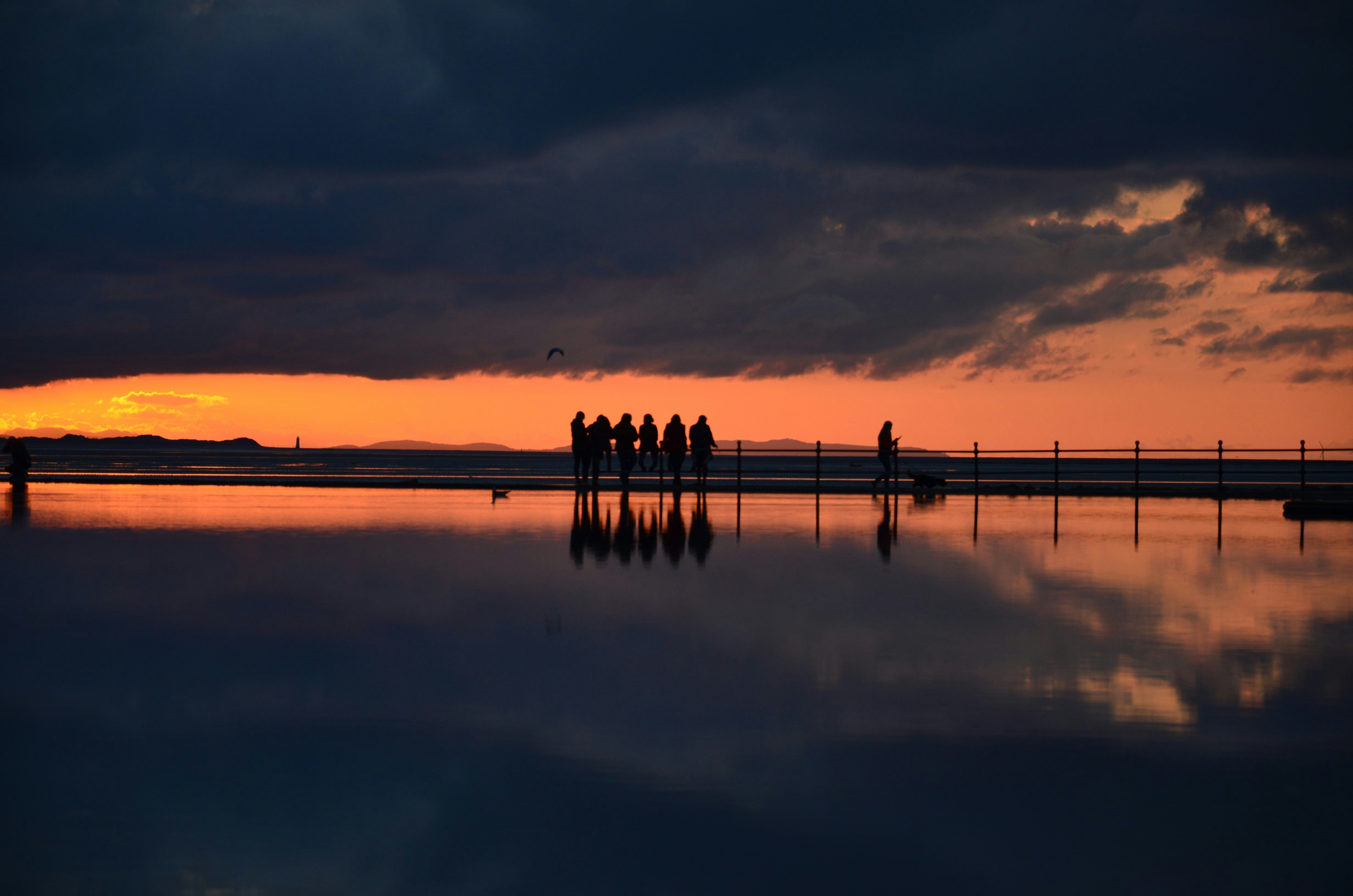 Silhouetted figures gather near the water's edge under a dramatic sunset sky.