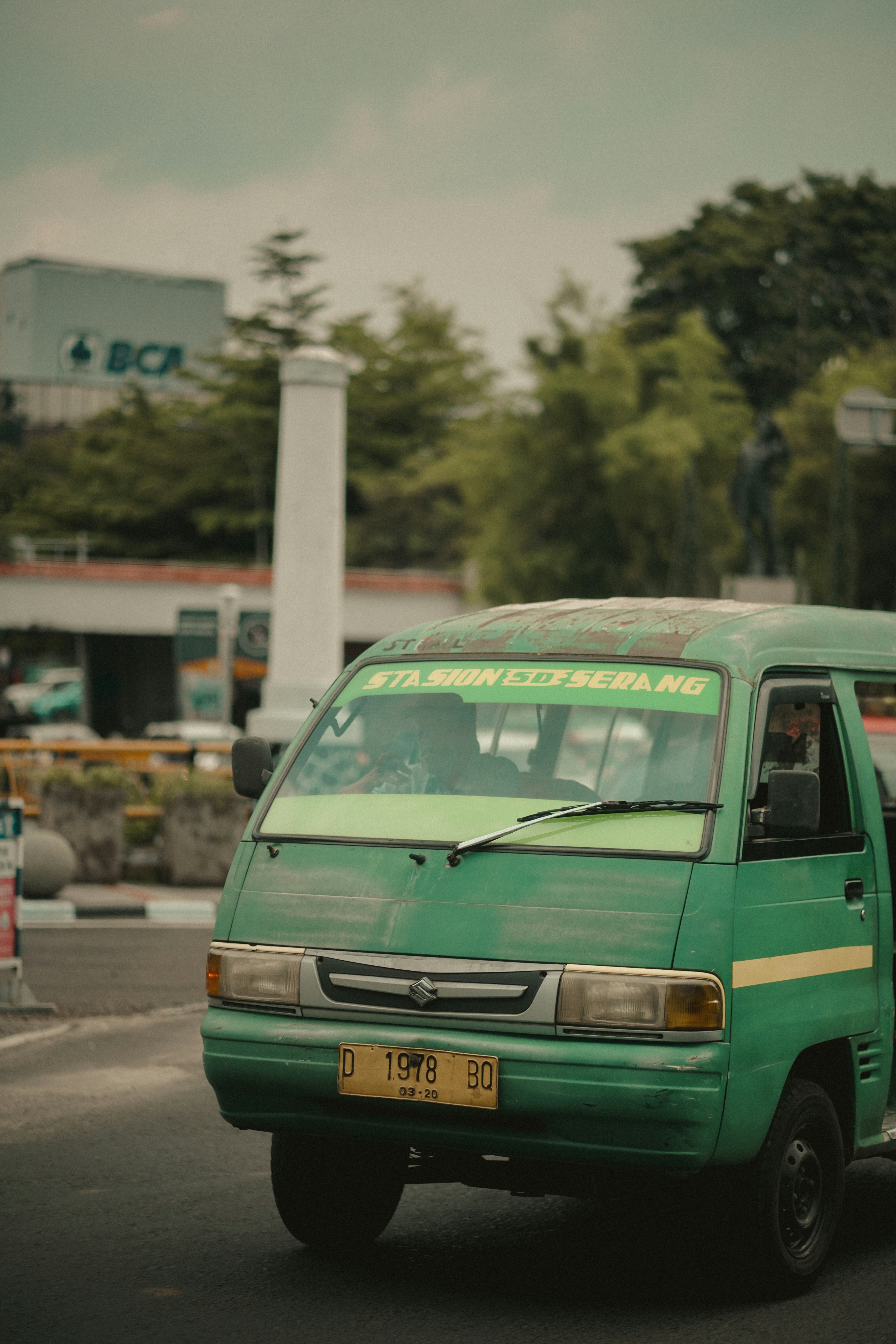 Green minivan with a faded sign indicating its route, navigating an urban intersection surrounded by trees and buildings.