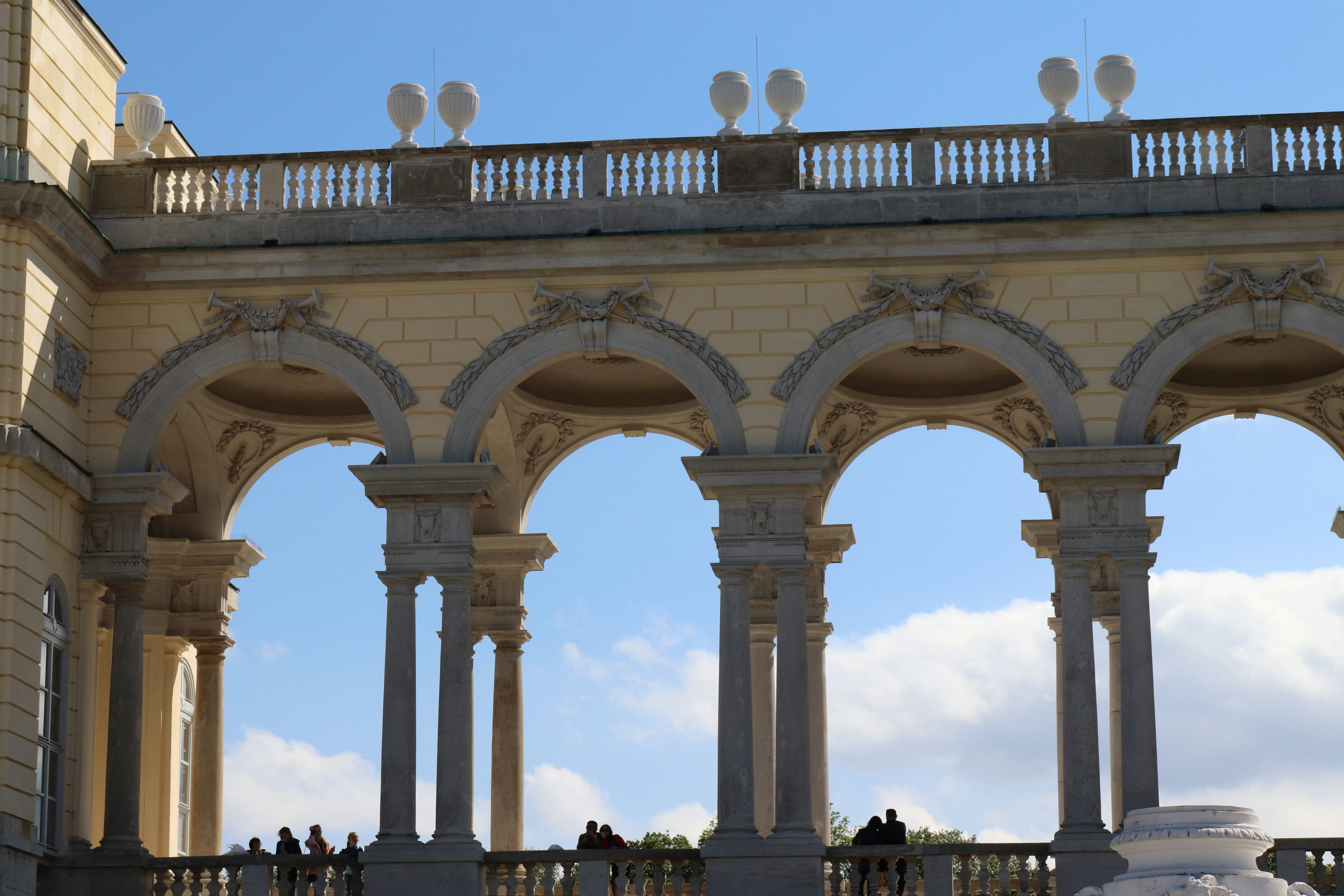Elegant arches of a historic structure frame visitors enjoying a sunny day, with a backdrop of soft clouds. The interplay of light and shadow enhances the architectural details.