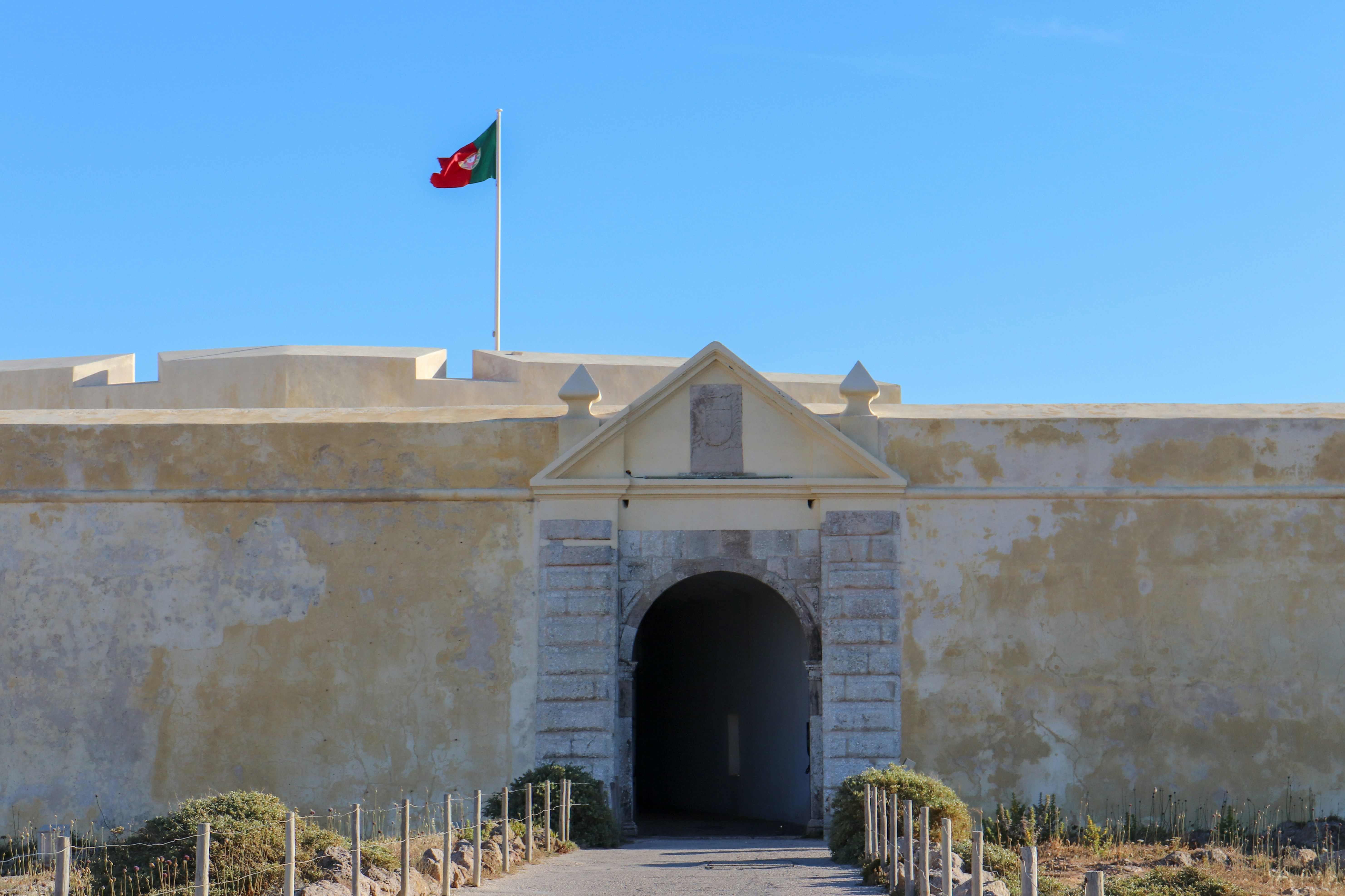 Historic fort entrance framed by weathered walls, featuring a flag fluttering above. The pathway leads into the fort's interior.