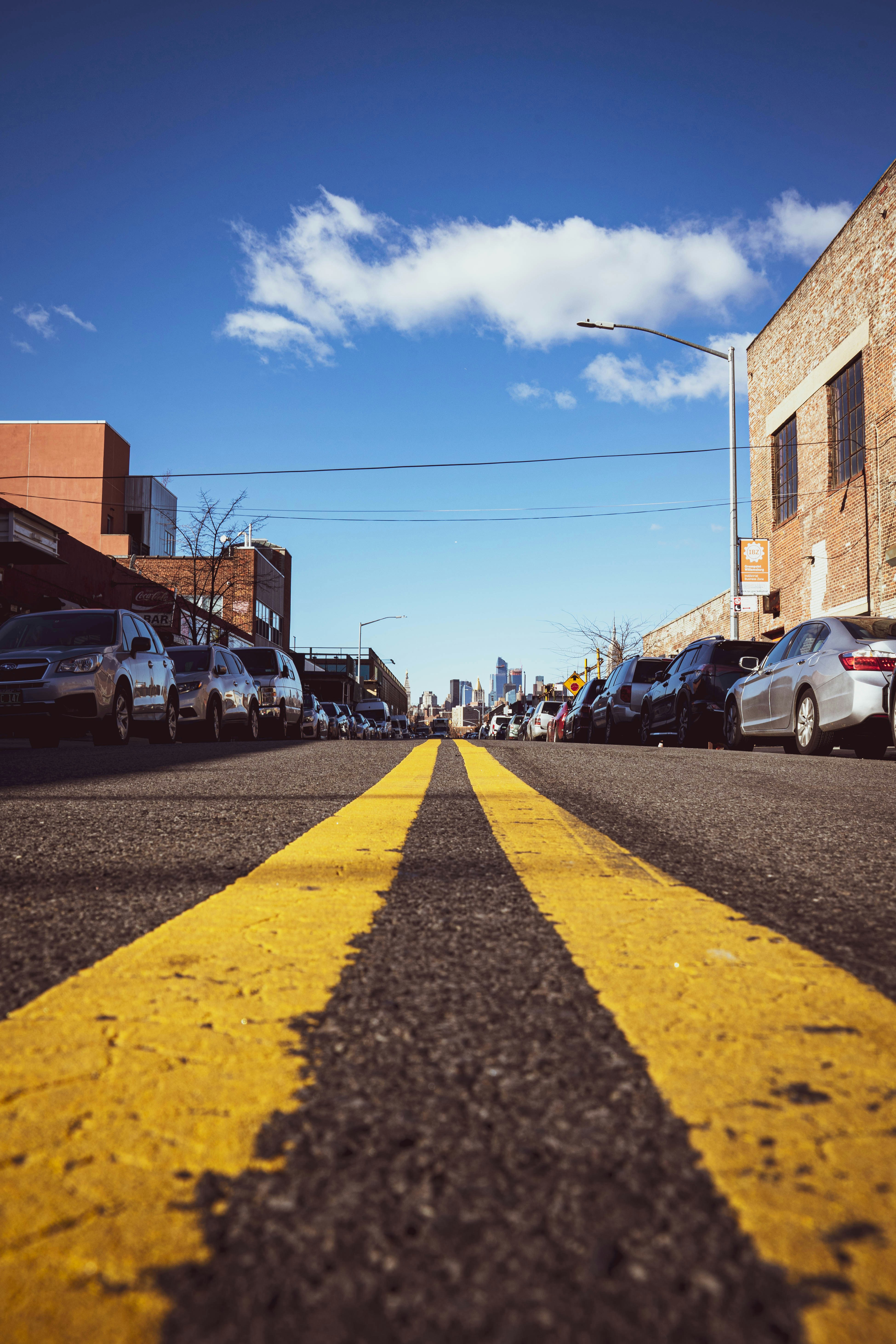 A low-angle view of a city street featuring yellow double lines and parked cars, leading towards a skyline in the distance.