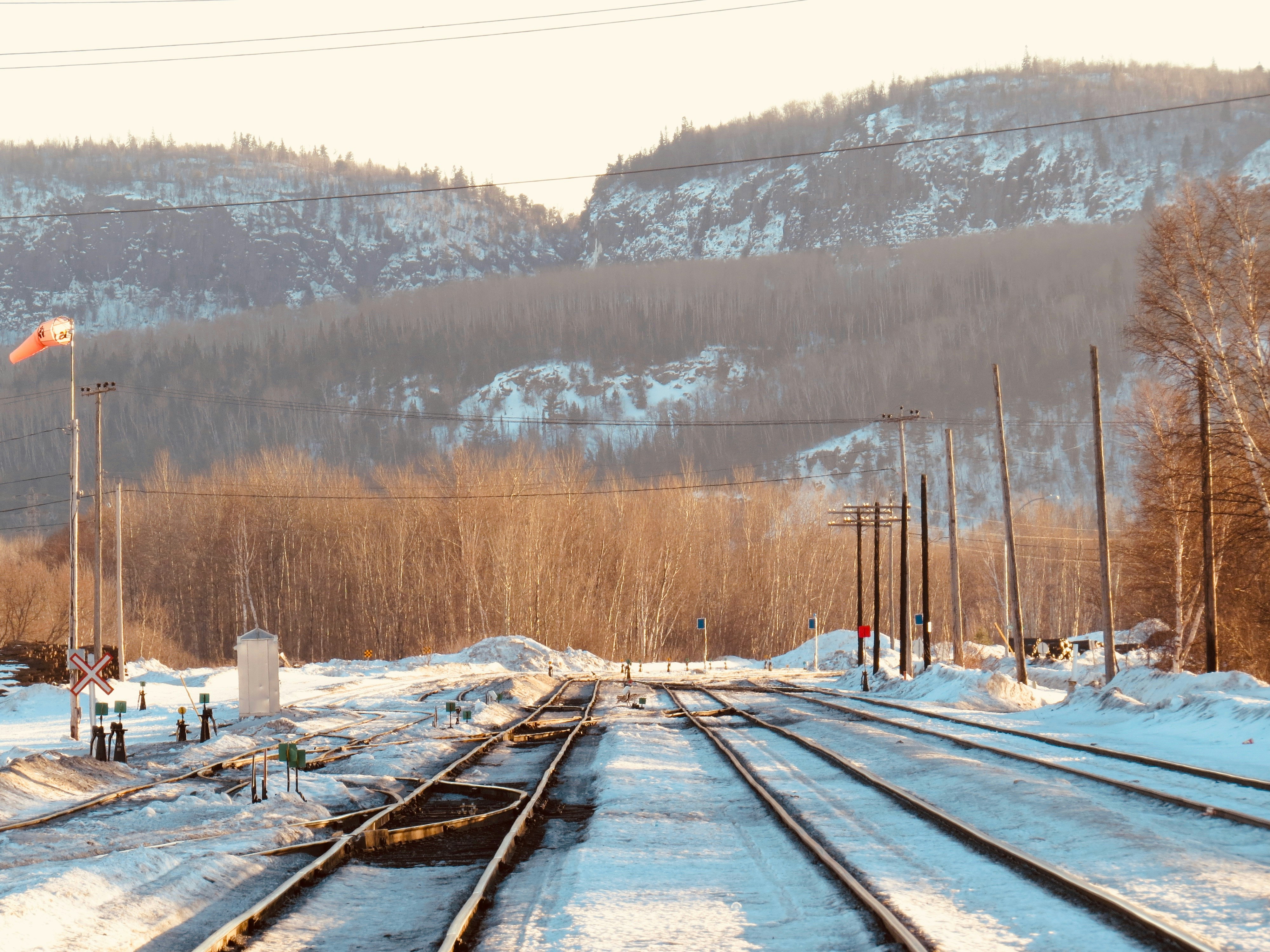 Snow-covered railway tracks leading towards distant mountains and a clear sky, with a hint of human activity in the foreground.