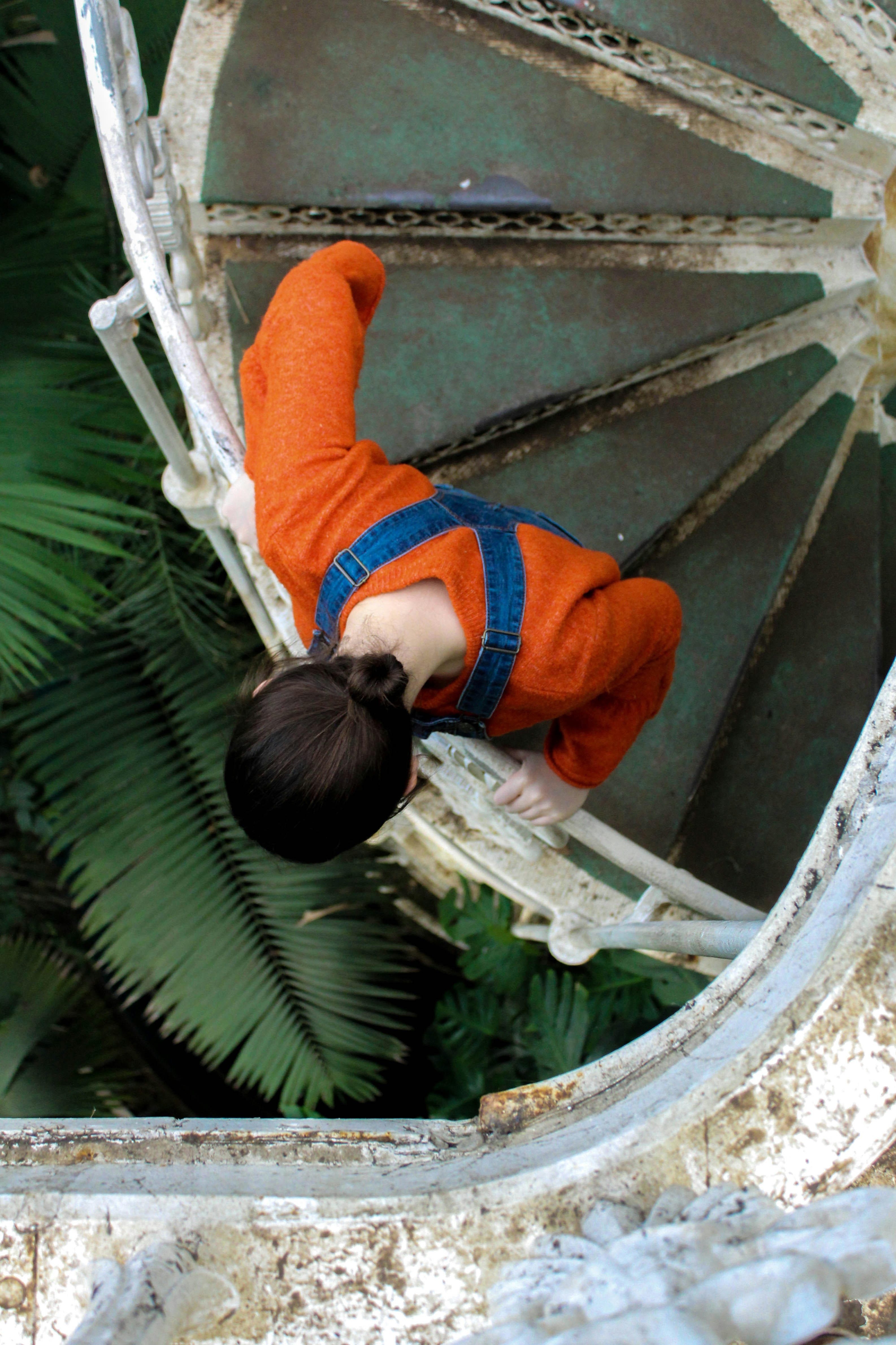 Spiral Descent: A child in orange overalls descends a weathered metal spiral staircase above tropical greenery