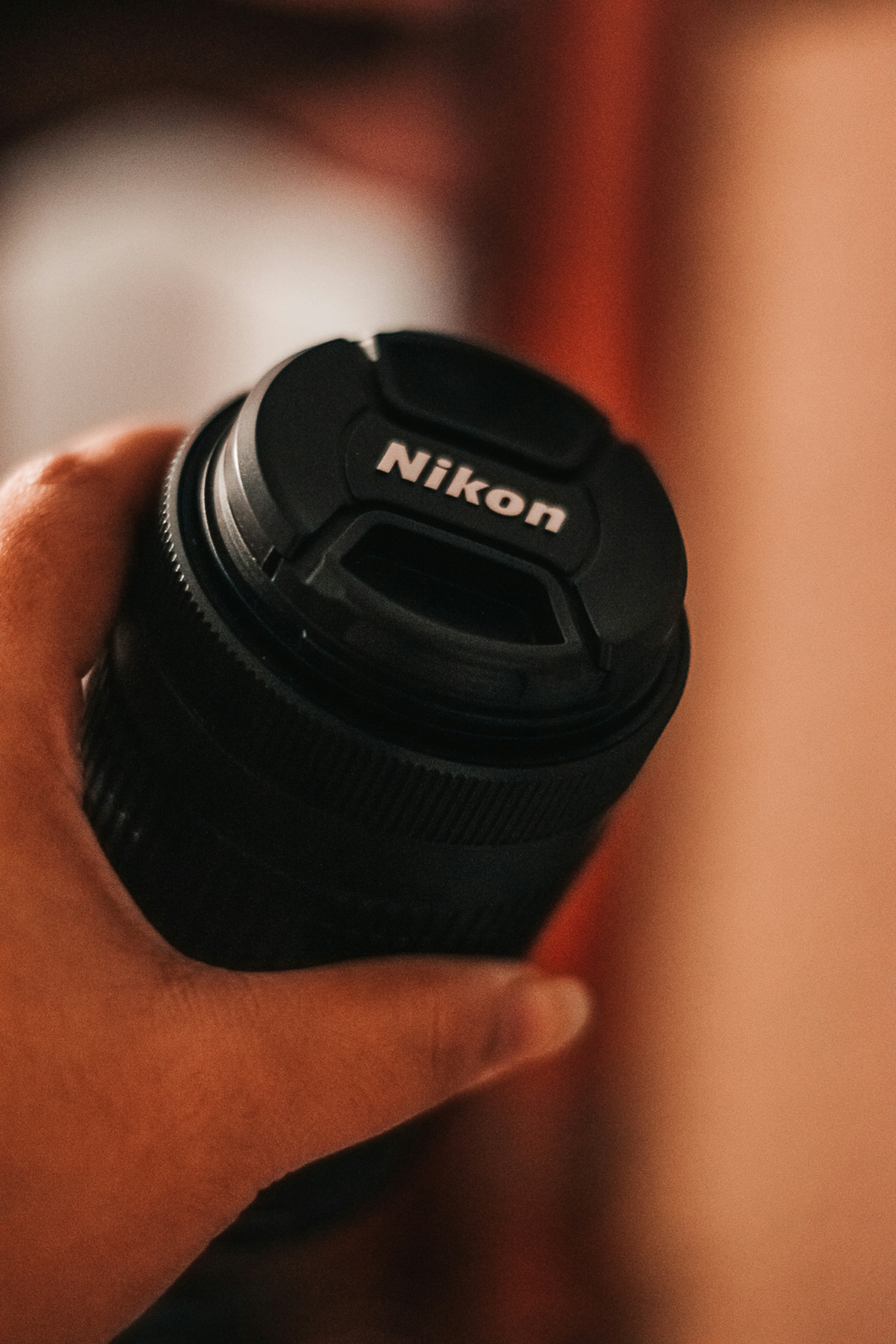 A hand holding a Nikon camera lens cap, showcasing the brand's signature design. The background features a soft bokeh effect that emphasizes the lens.