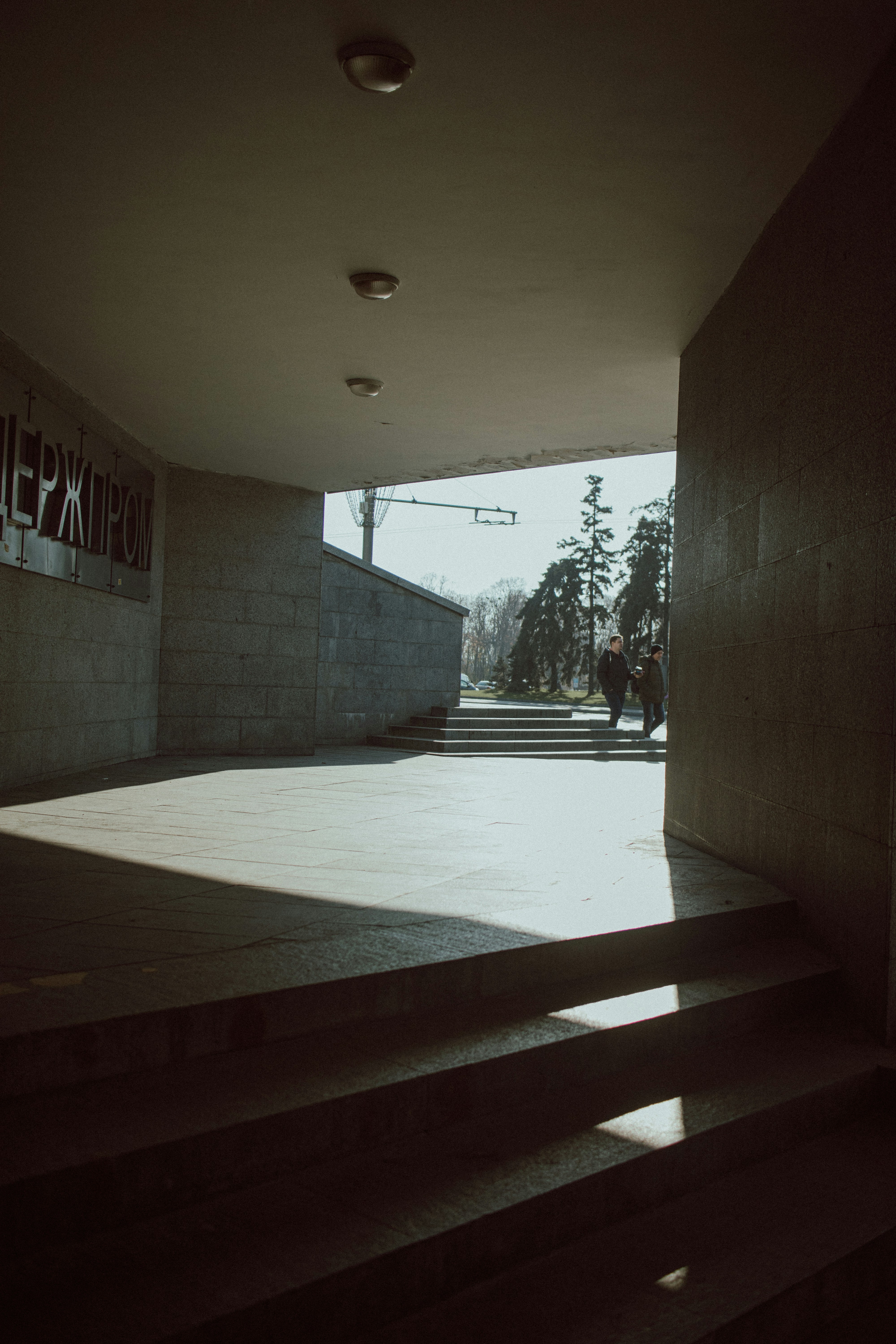 Sunlight filters through an archway, illuminating a staircase leading to a park where two figures walk. The scene captures a moment of transition and exploration.