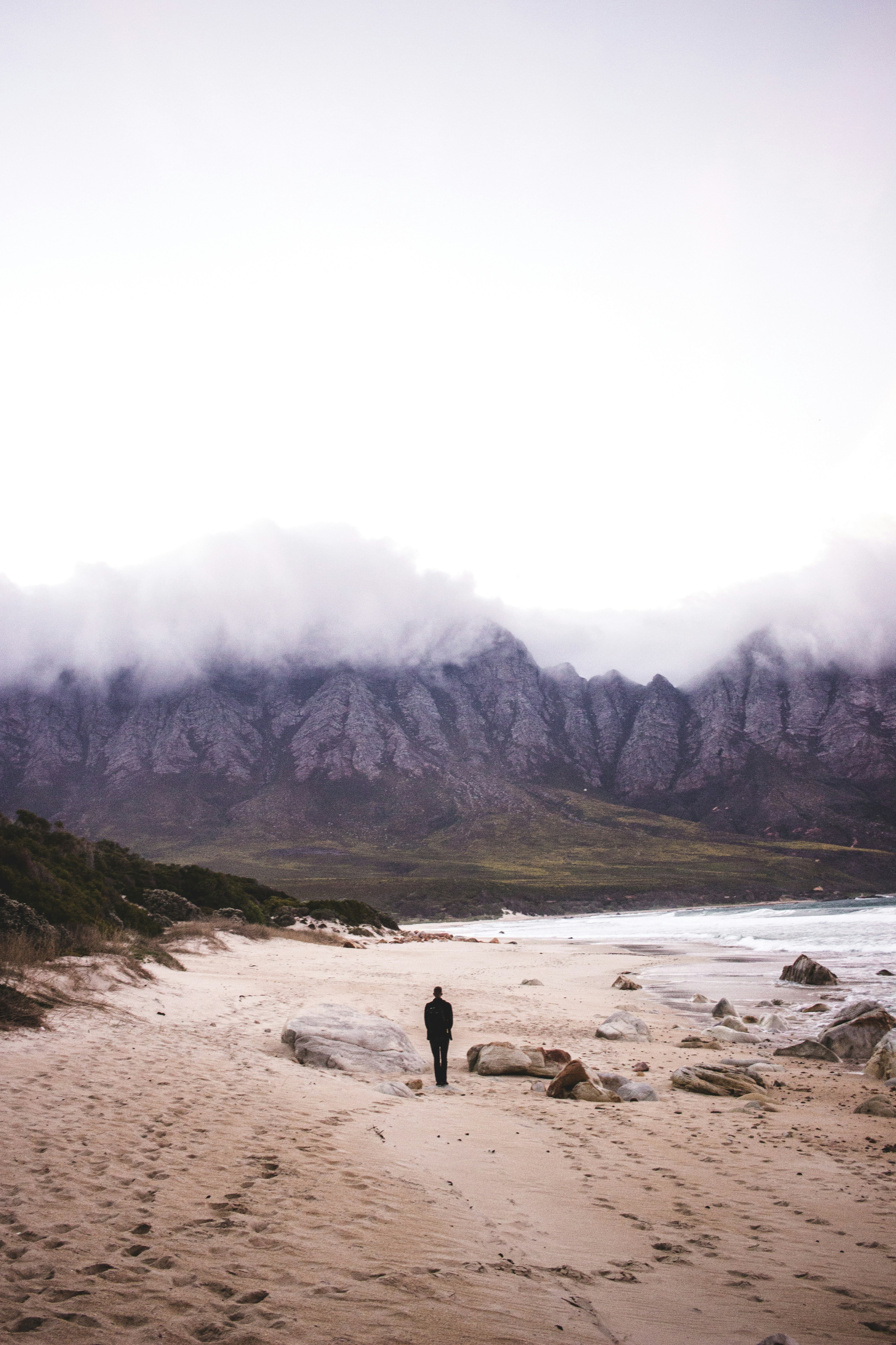 A solitary figure stands on a sandy beach, framed by dramatic mountains shrouded in mist. The scene captures a moment of reflection amidst nature's grandeur.