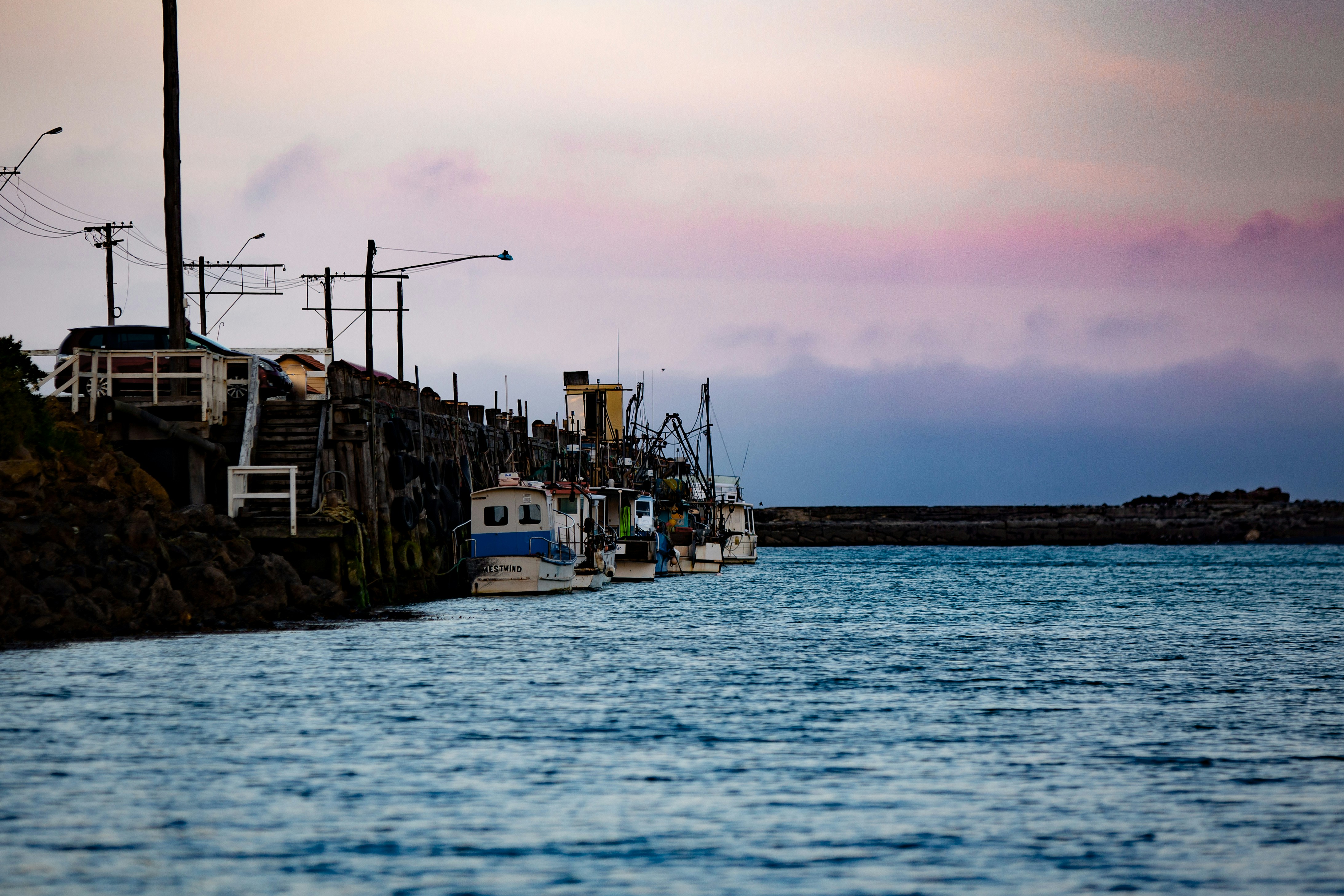 Fishing boats lined up along a tranquil harbor at dusk, with soft pastel skies reflecting on the water's surface.