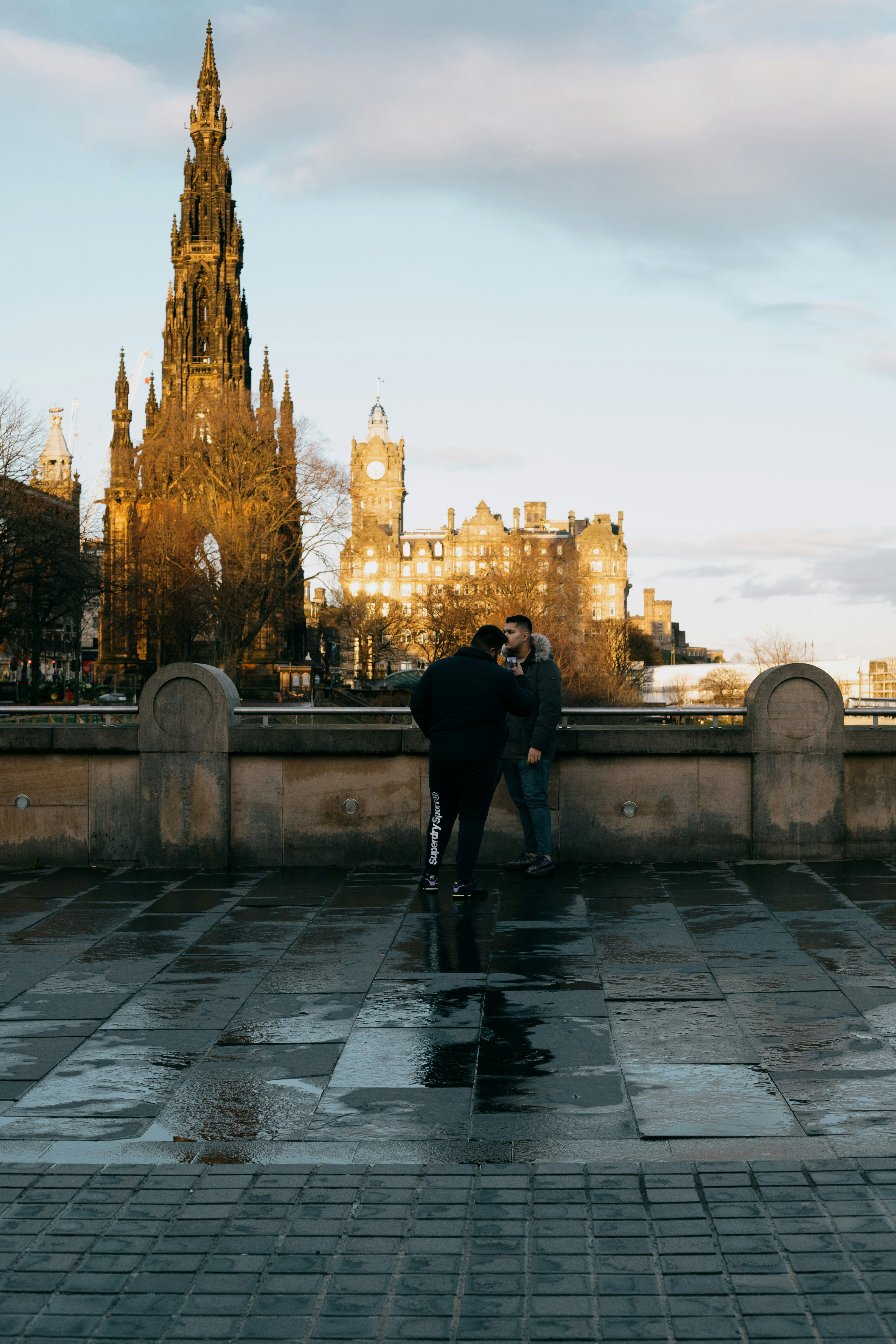 Two men share an intimate conversation on a wet stone pavement, with the iconic Scott Monument and historical buildings illuminated in the background.