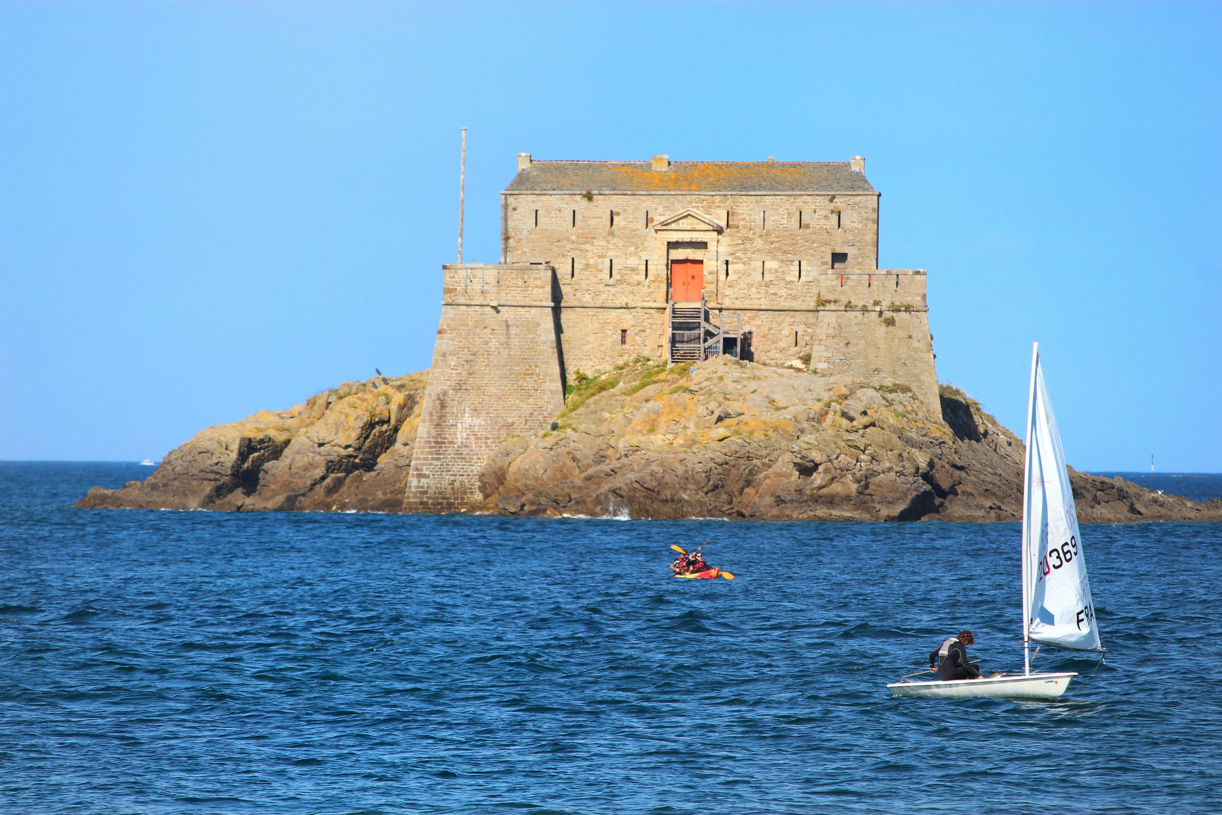 Stone fortress perched on a rocky island surrounded by sailboats on a clear blue sea.