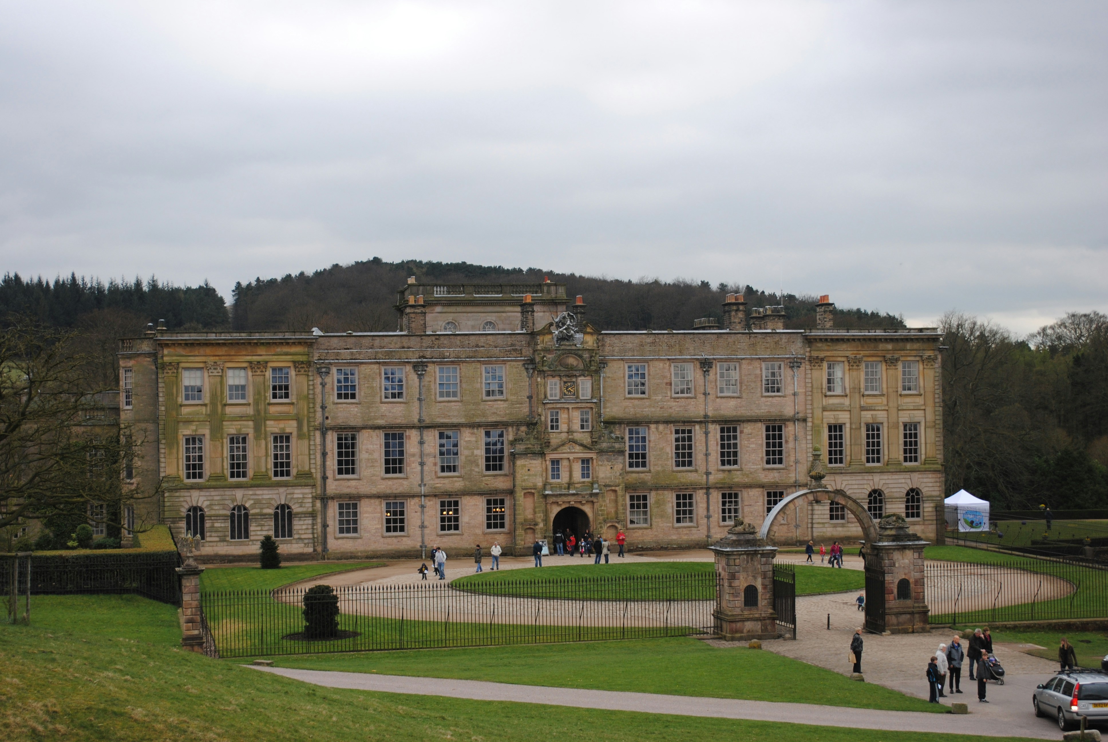 Grand stone mansion with symmetrical architecture set against a backdrop of lush hills and cloudy sky.