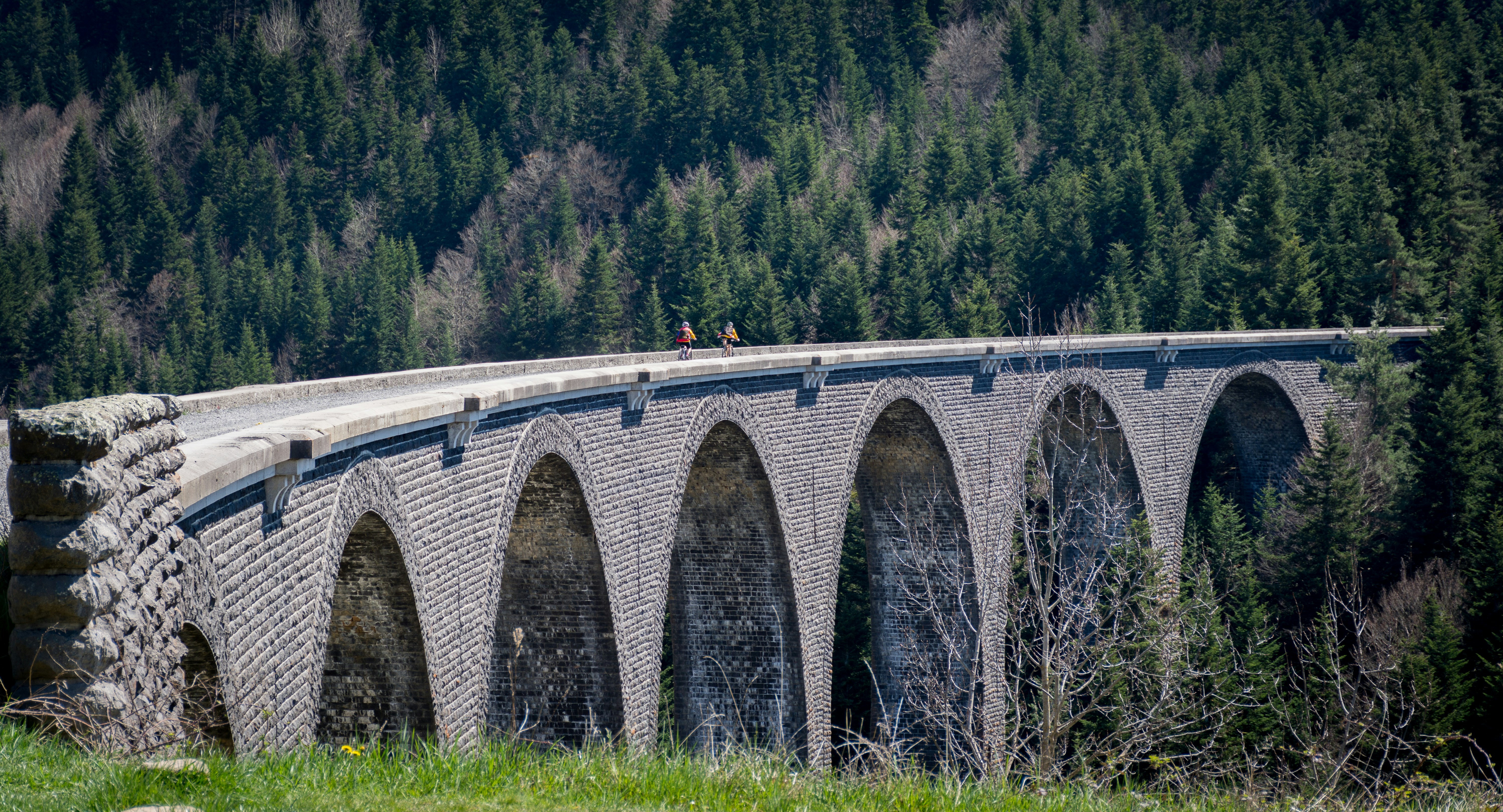 Historic stone viaduct spanning a lush forested valley, with hikers visible on the bridge. The structure showcases remarkable architectural design.