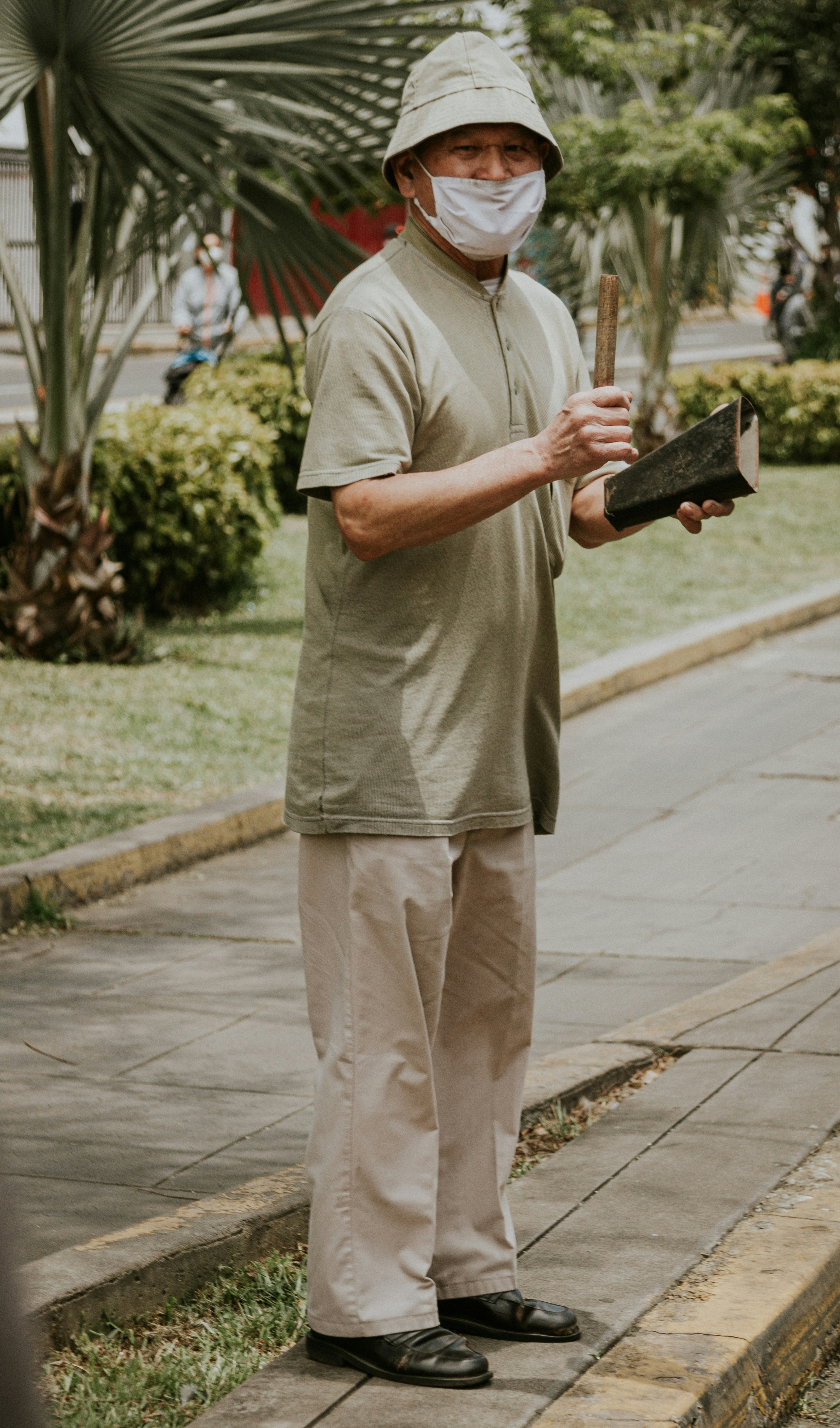 Elderly man holding a wooden tool and a book, standing on a city sidewalk surrounded by greenery.