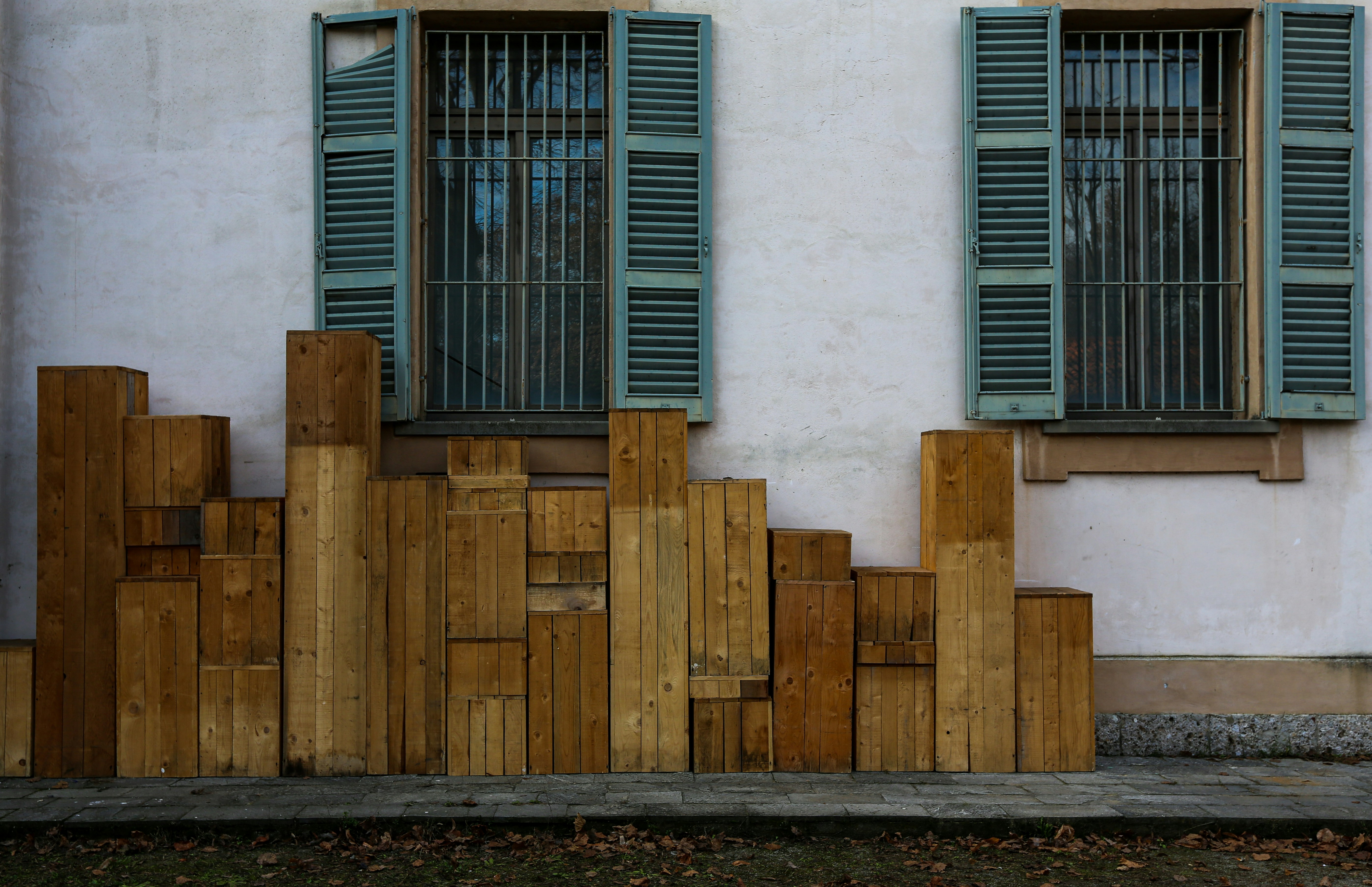 Wooden structures arranged in varying heights against a pastel wall with green-shuttered windows. A blend of natural and architectural elements creates an intriguing composition.