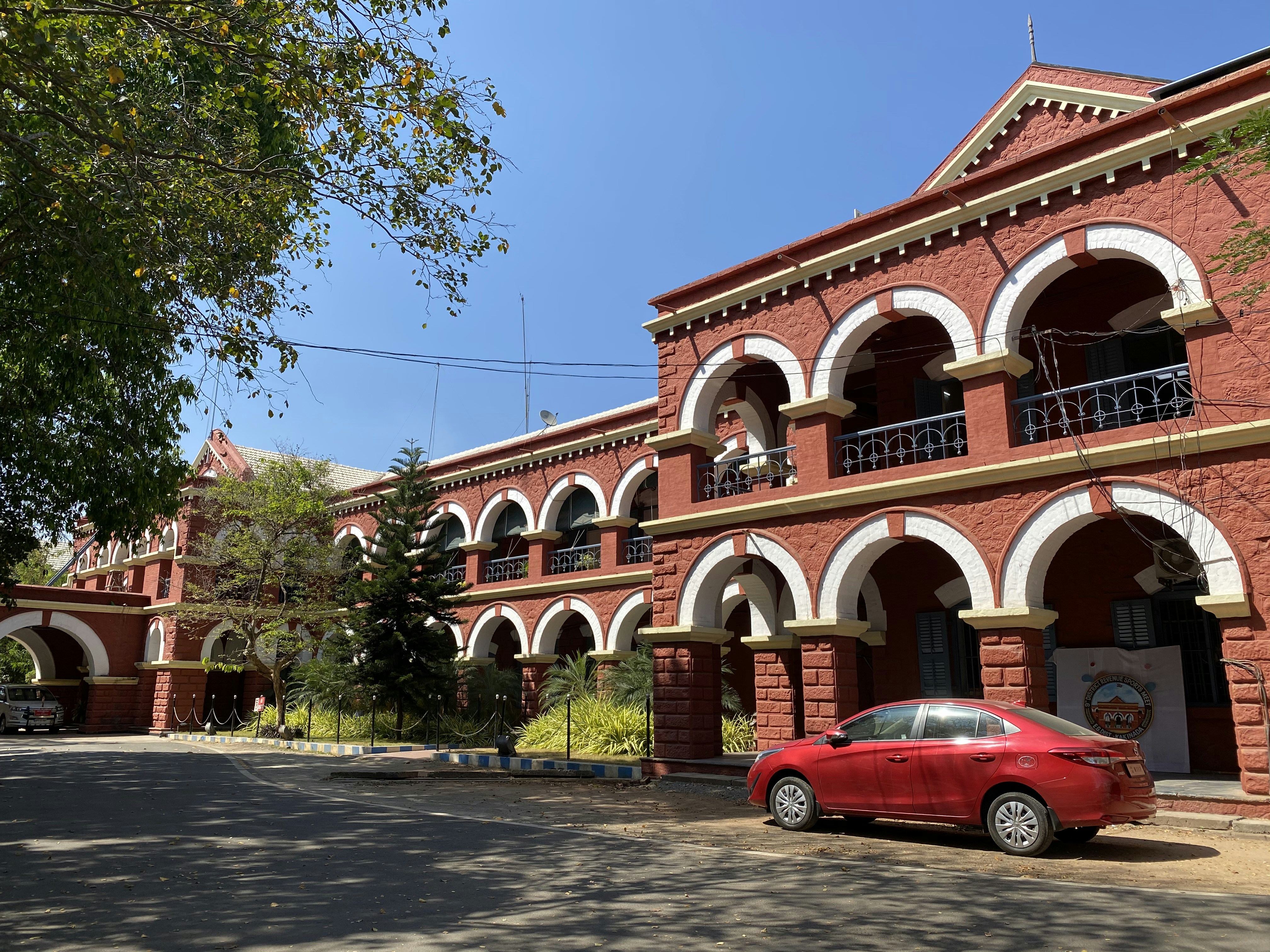 Red car parked in front of a historic red-brick building with arched windows under a clear blue sky.