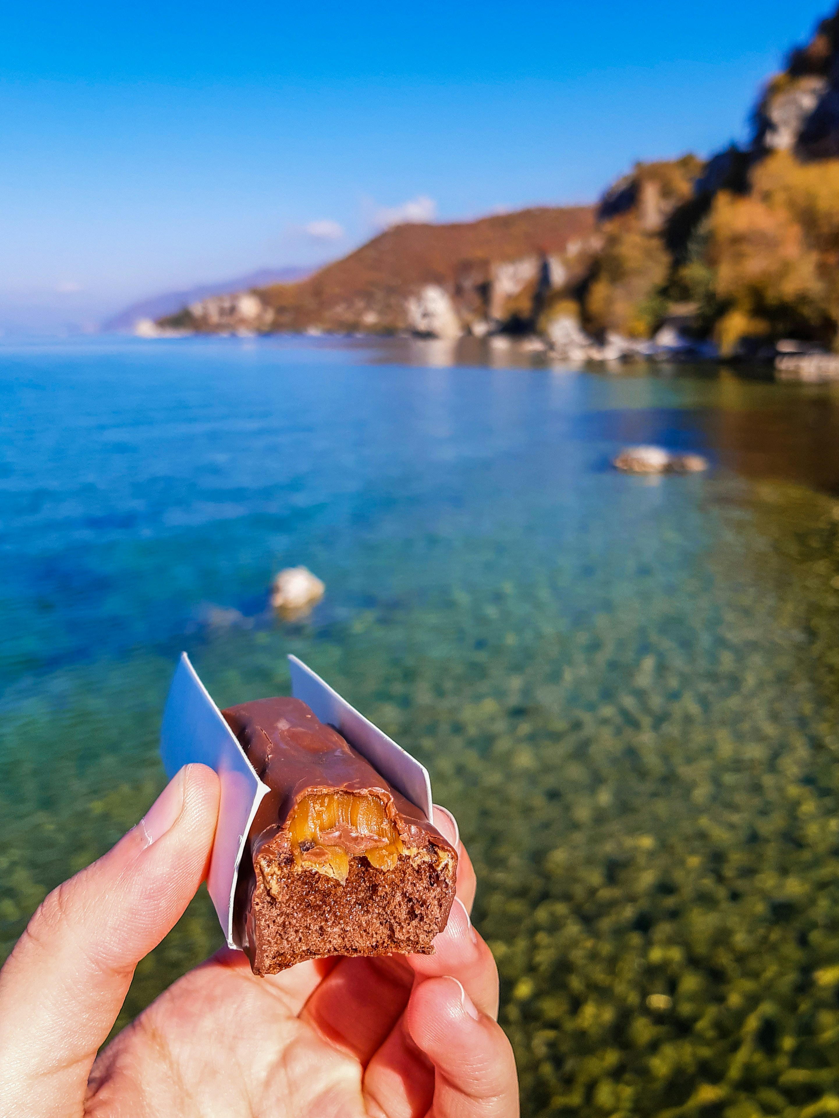 Handheld chocolate pastry in a white wrapper is held above a turquoise alpine lake, with the distant shoreline and hills appearing softly blurred.
