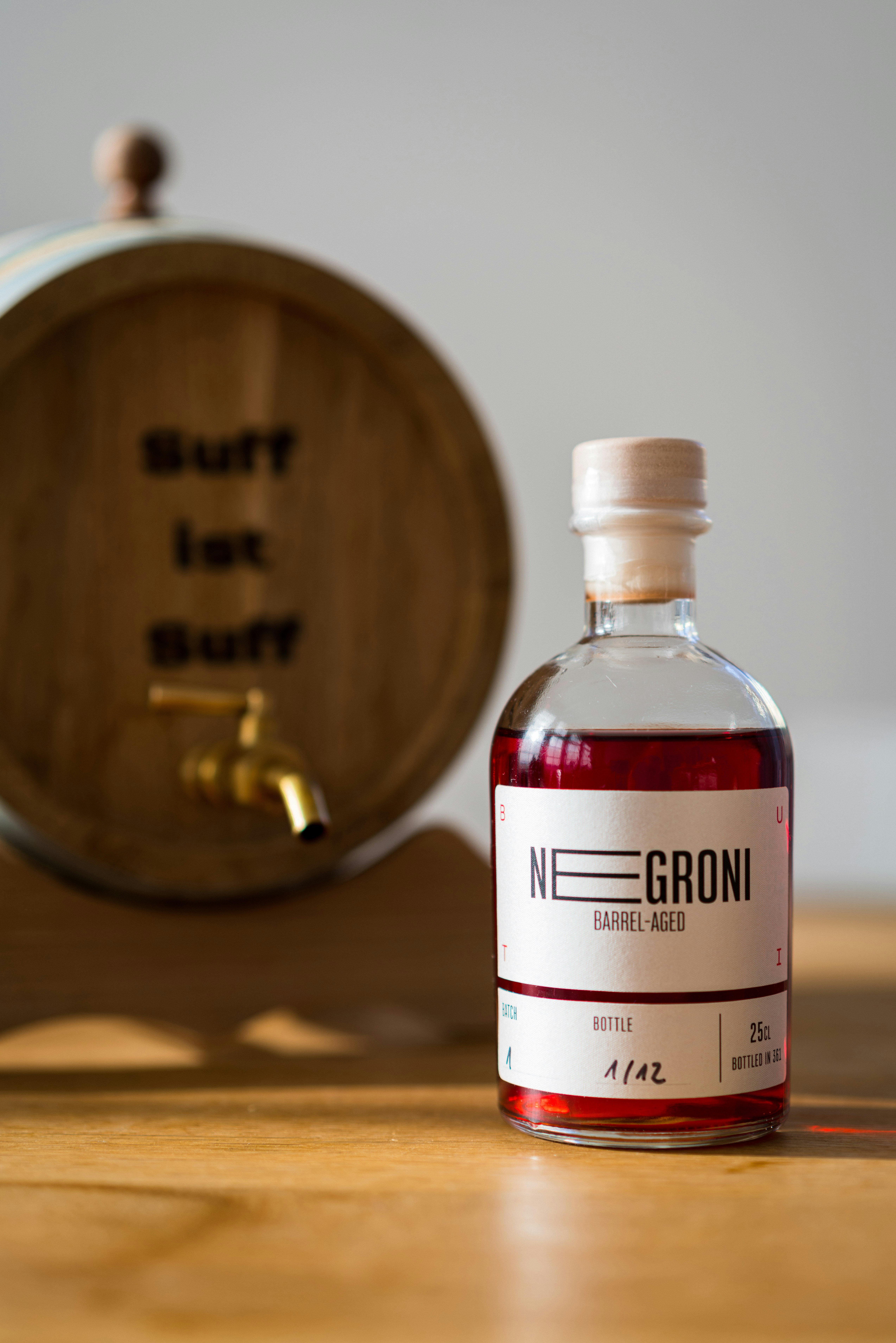 A bottle of barrel-aged Negroni sits elegantly on a wooden surface, with a rustic barrel in the background featuring playful text.