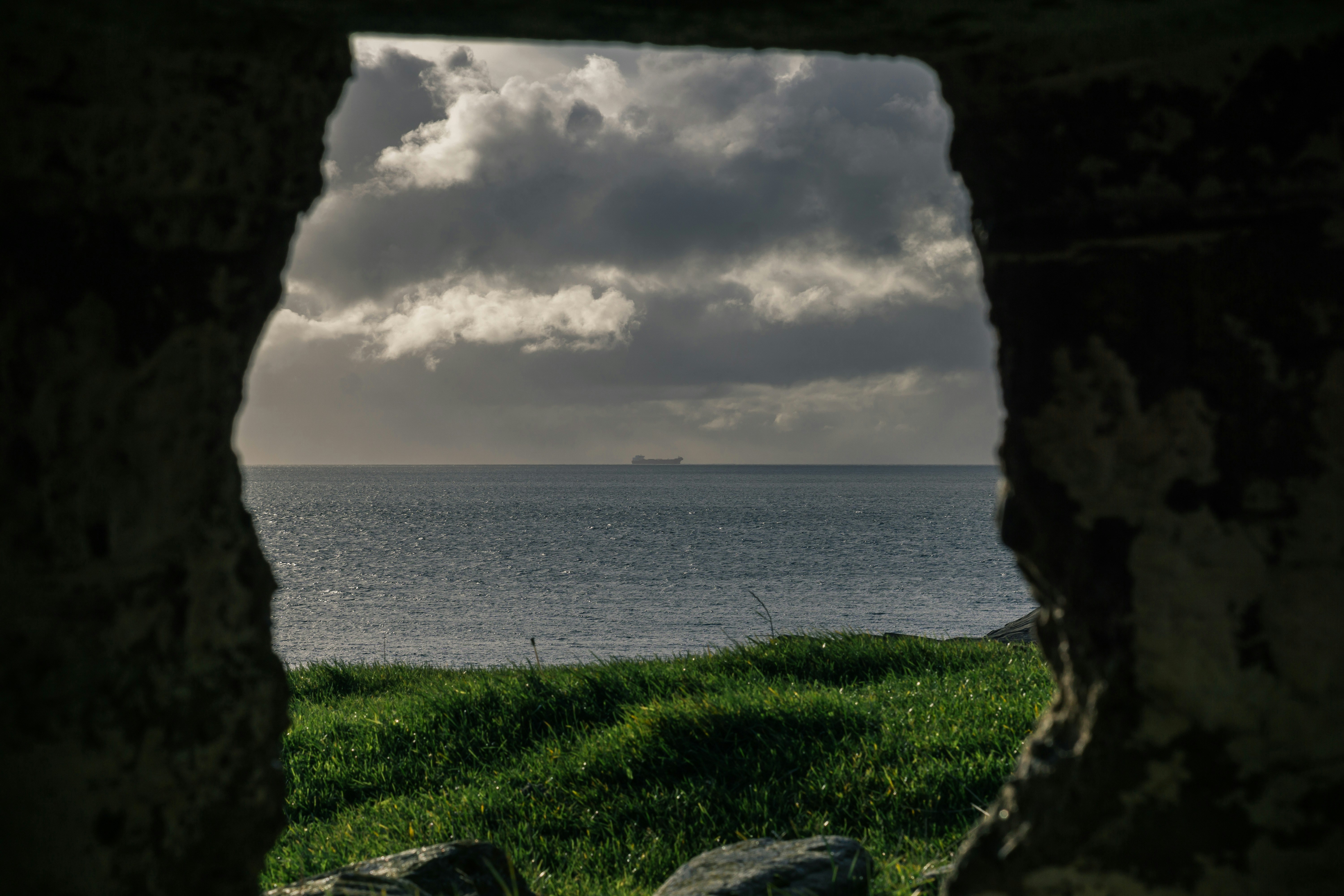View of a distant ship on the horizon framed by a weathered stone opening, contrasting the lush green grass below and the moody sky above.