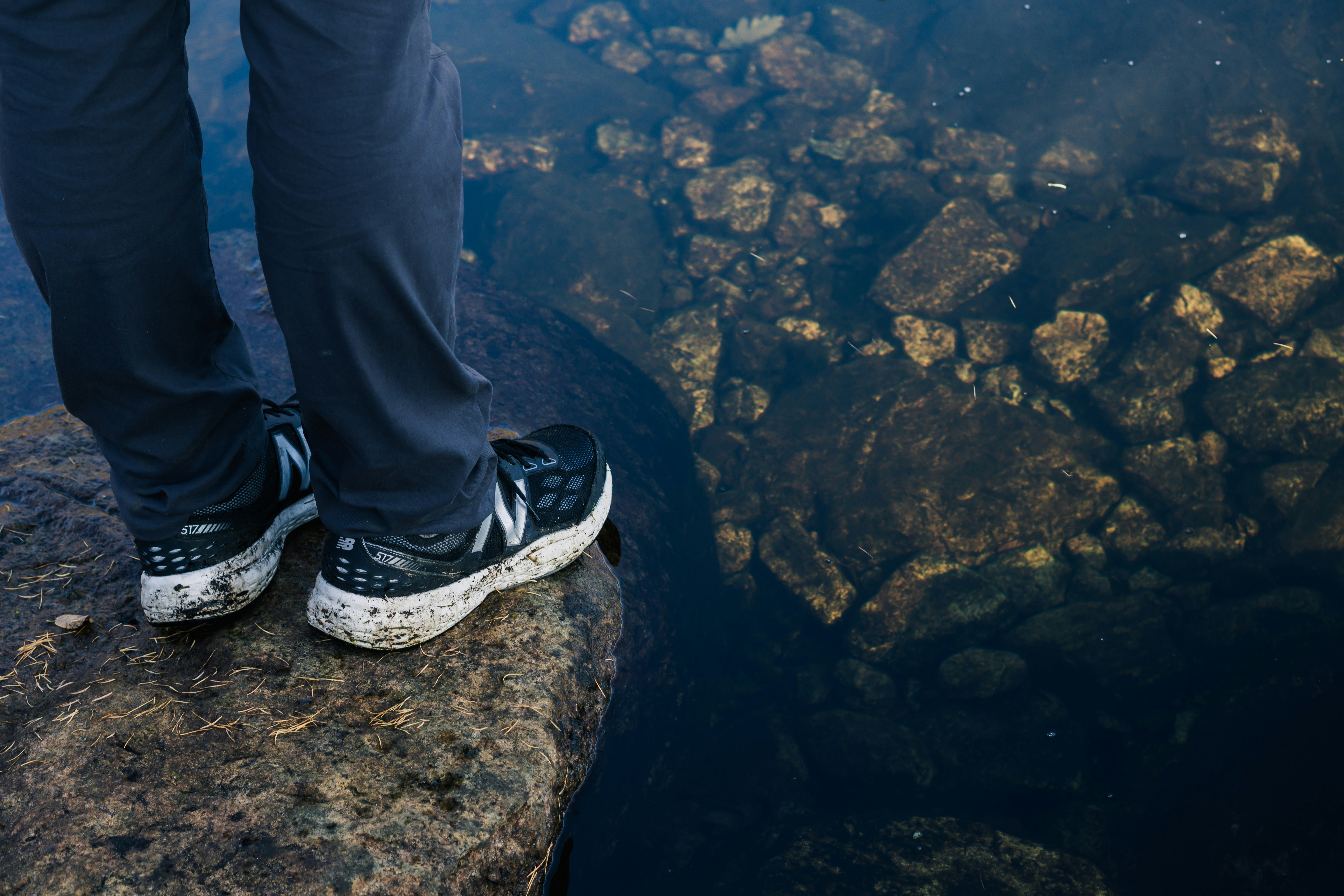 Person standing on a rocky ledge above clear water, with shoes partially submerged, creating a tranquil scene of nature's harmony.