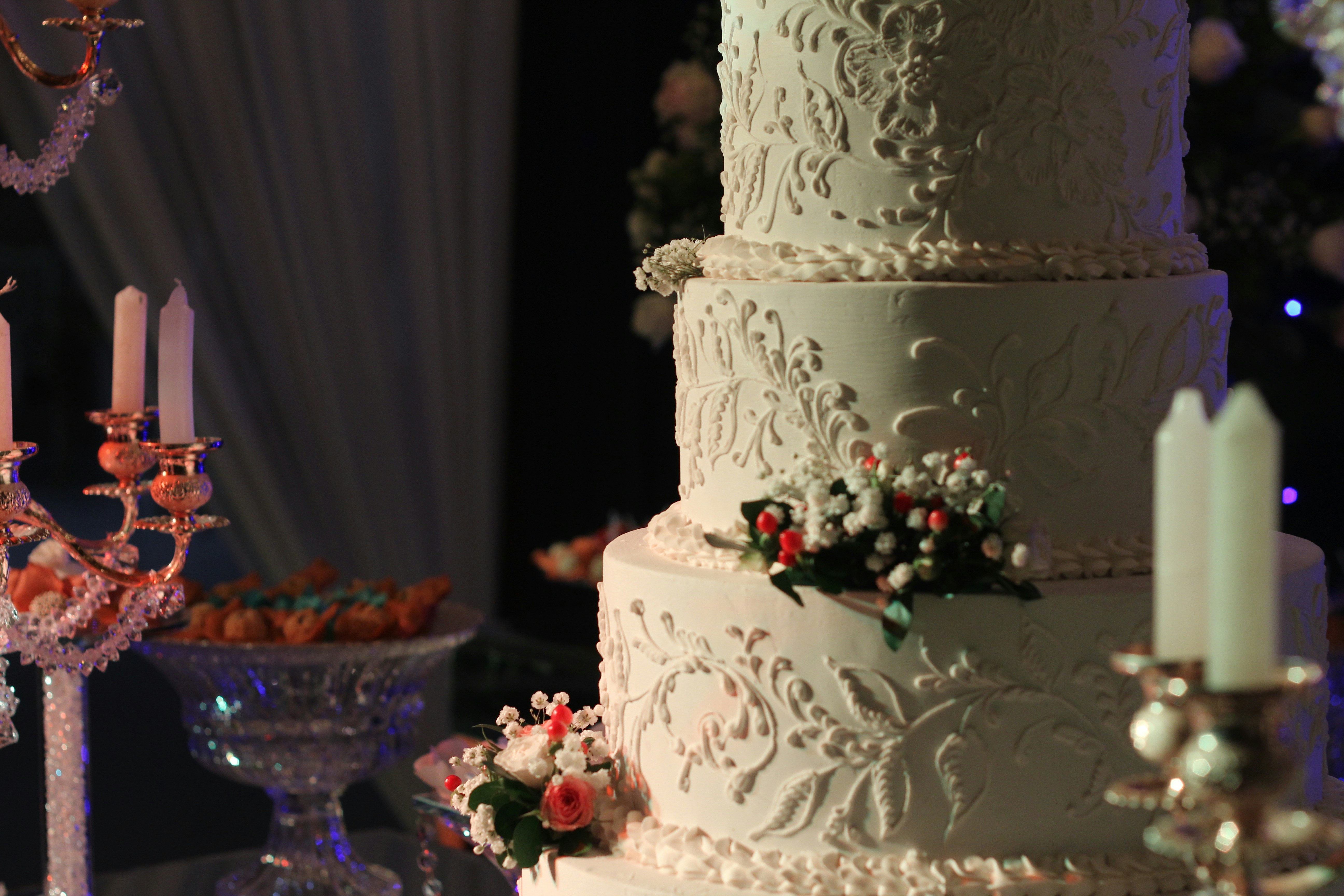 Intricately designed three-tier wedding cake adorned with floral accents, complemented by elegant candle holders and a dessert display in the background.