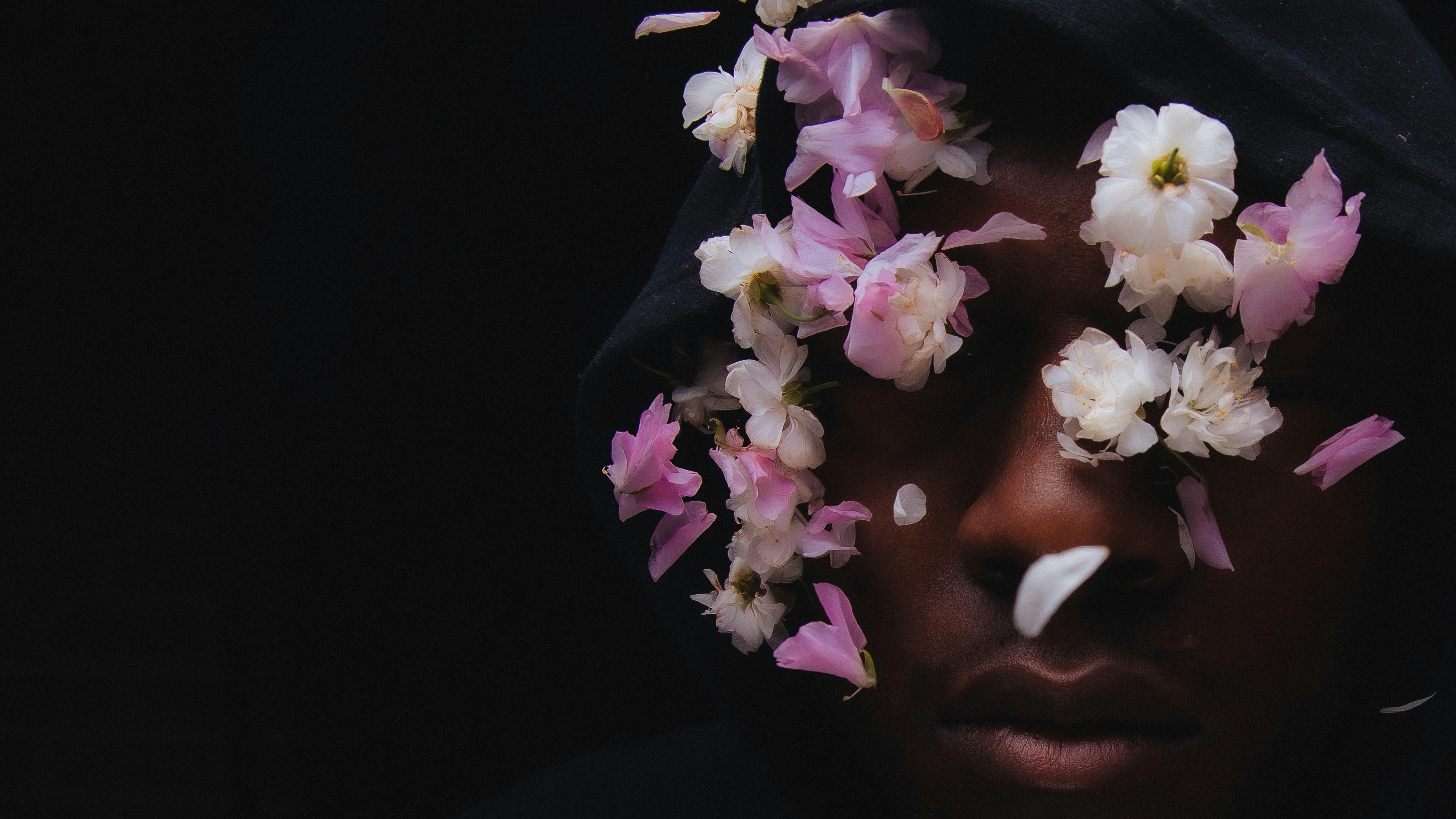 A portrait featuring a person with flowers delicately arranged across their face, set against a dark background. The contrast highlights the intricate details of the blossoms.