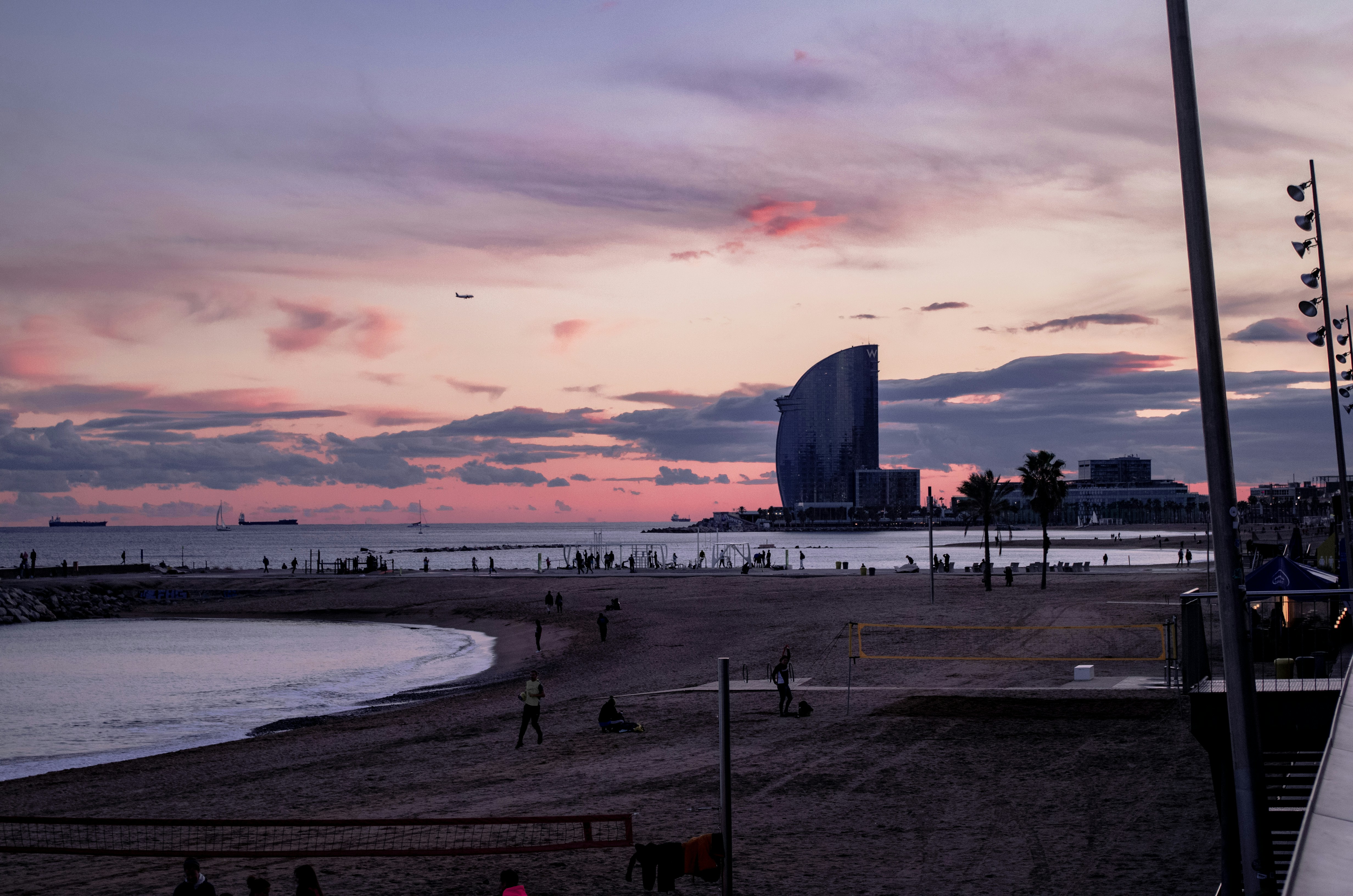 Silhouetted figures stroll along the beach as the sun sets behind a modern skyline, casting a warm glow across the water. 