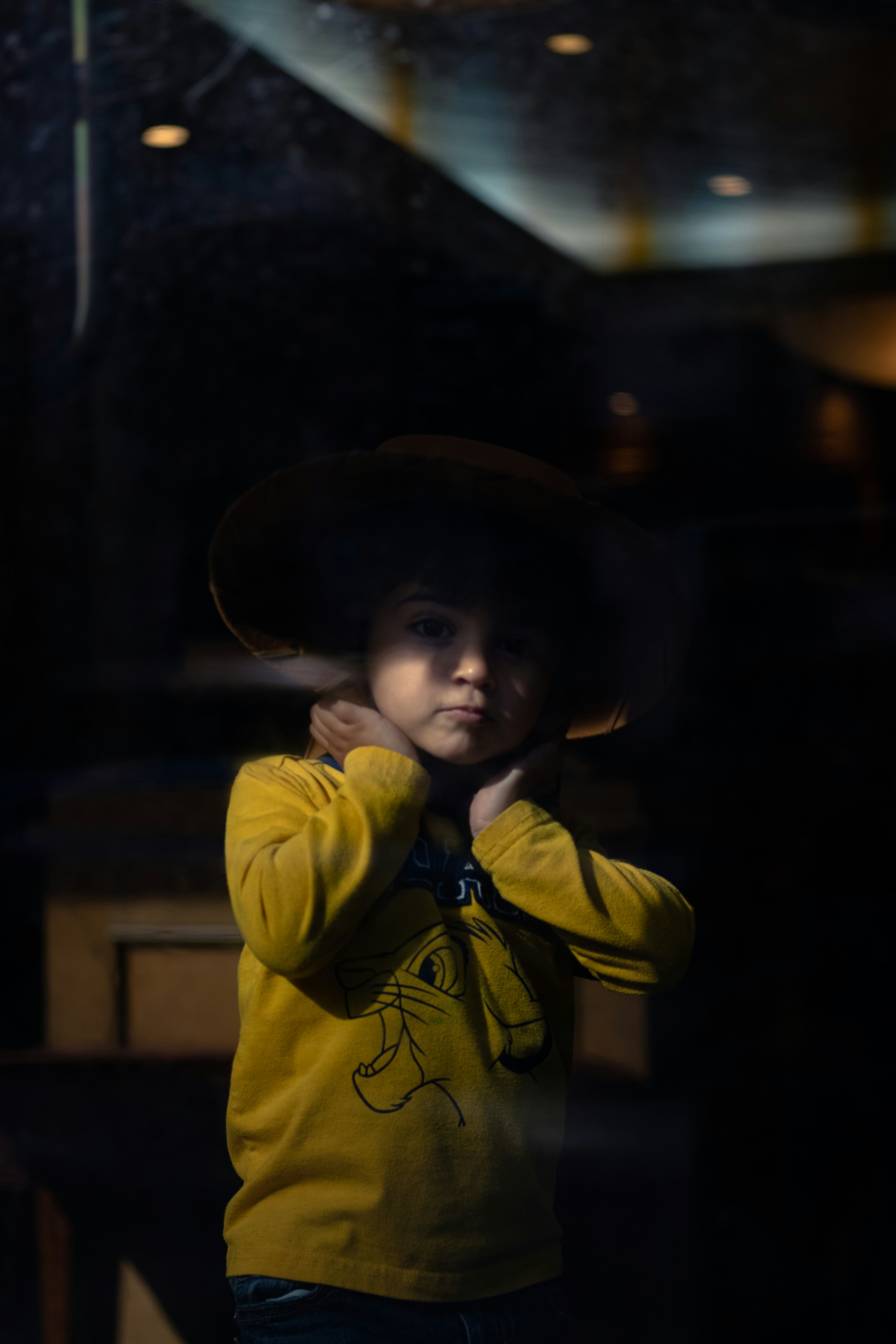 A young child in a yellow sweater and wide-brimmed hat stands quietly, framed by soft shadows and light reflections. The atmosphere evokes a sense of innocence and contemplation.