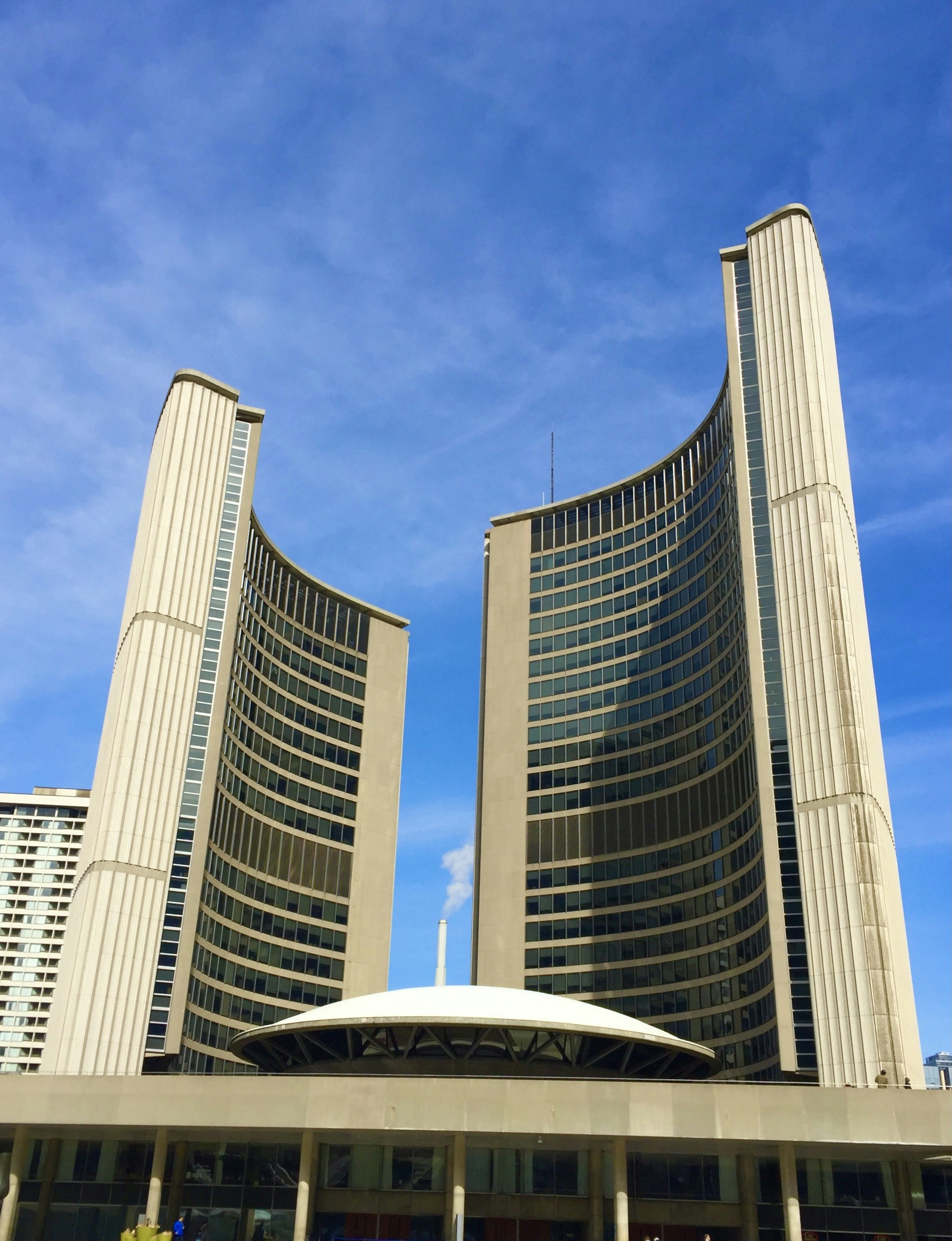 Two towering architectural structures with curved facades rise against a clear blue sky, showcasing modern design elements. A circular canopy sits at the base, adding to the contemporary aesthetic.