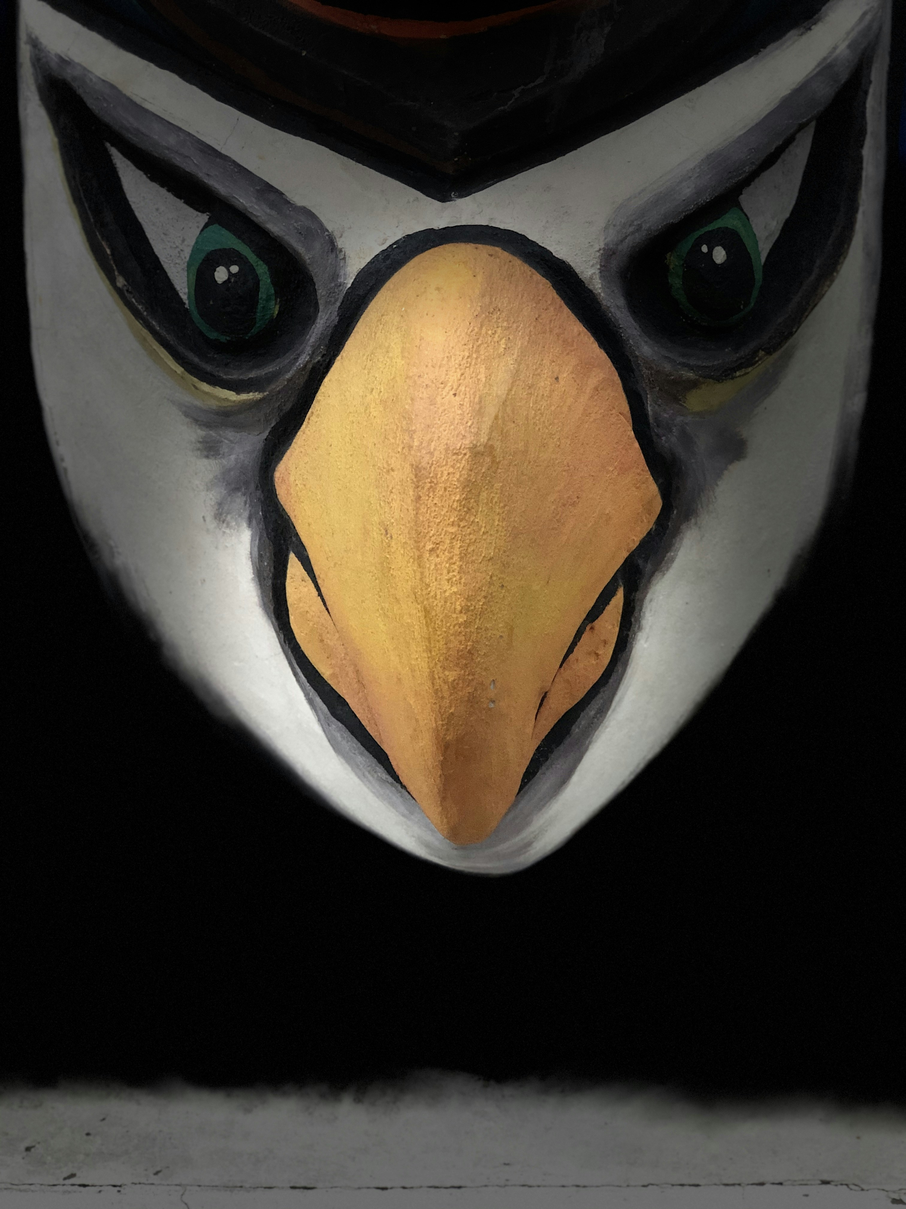 A vividly painted bird mask with piercing eyes and a prominent beak, set against a dark background.