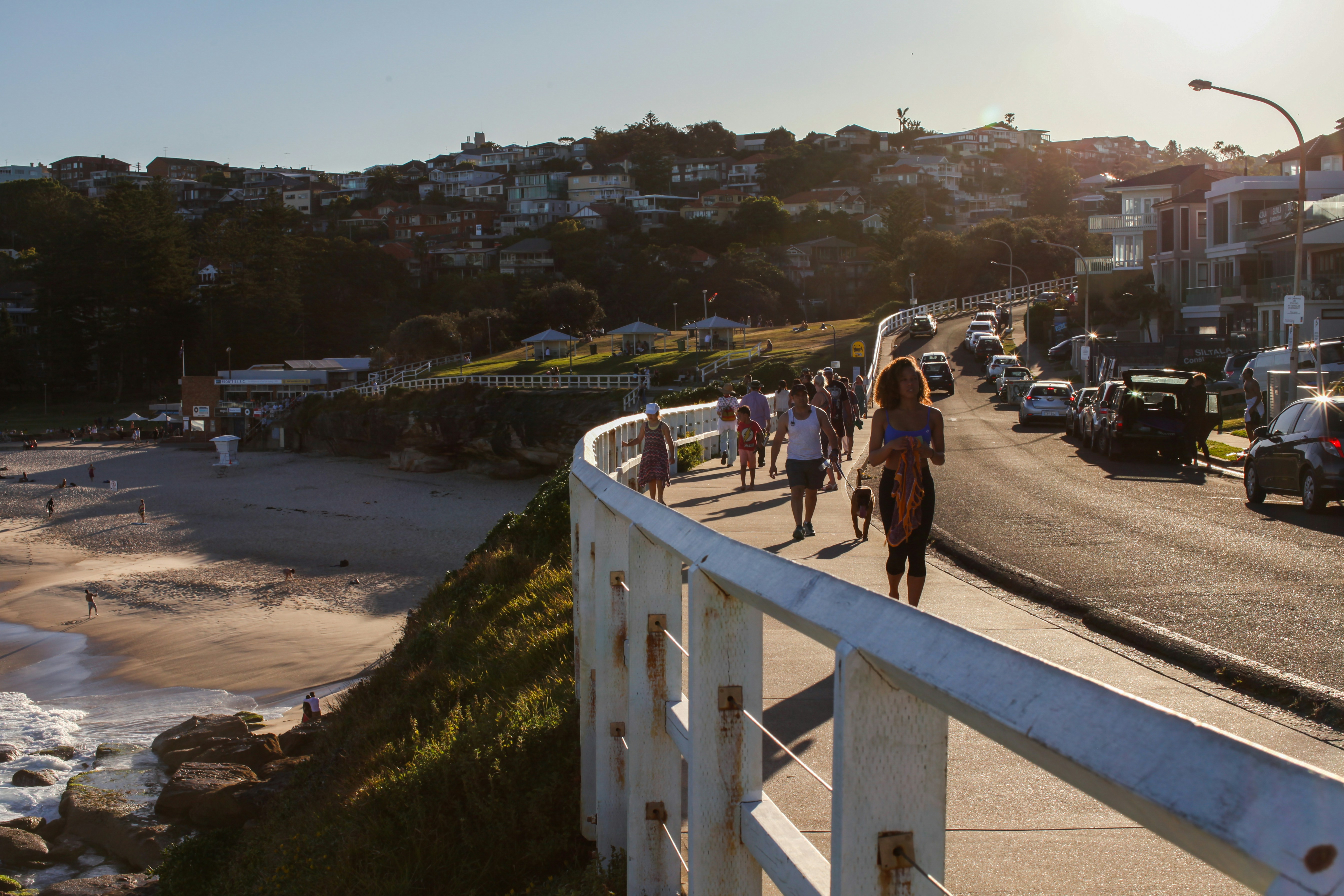 A lively coastal walkway with pedestrians enjoying the sunset, overlooking the beach and nearby houses. The scene captures the essence of a vibrant seaside community.