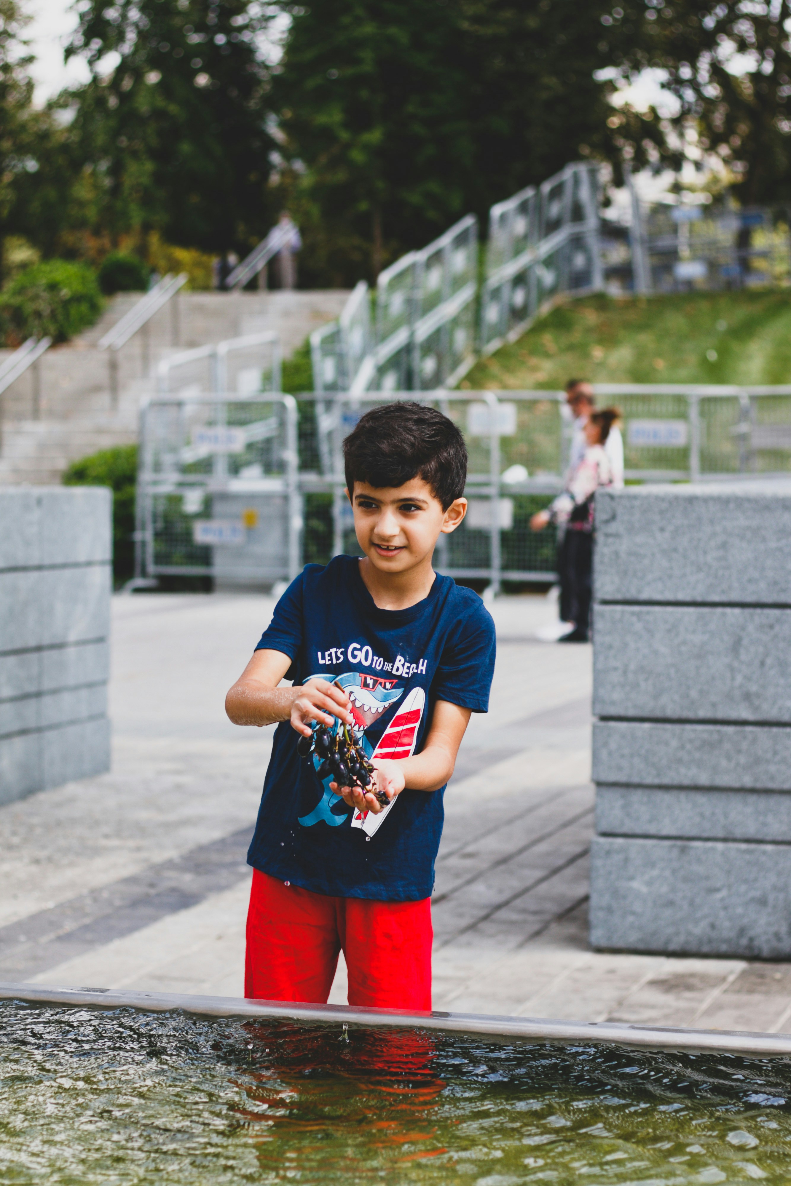 Young boy playfully splashing water while holding colorful toys at a public fountain. Bright surroundings enhance the cheerful atmosphere.