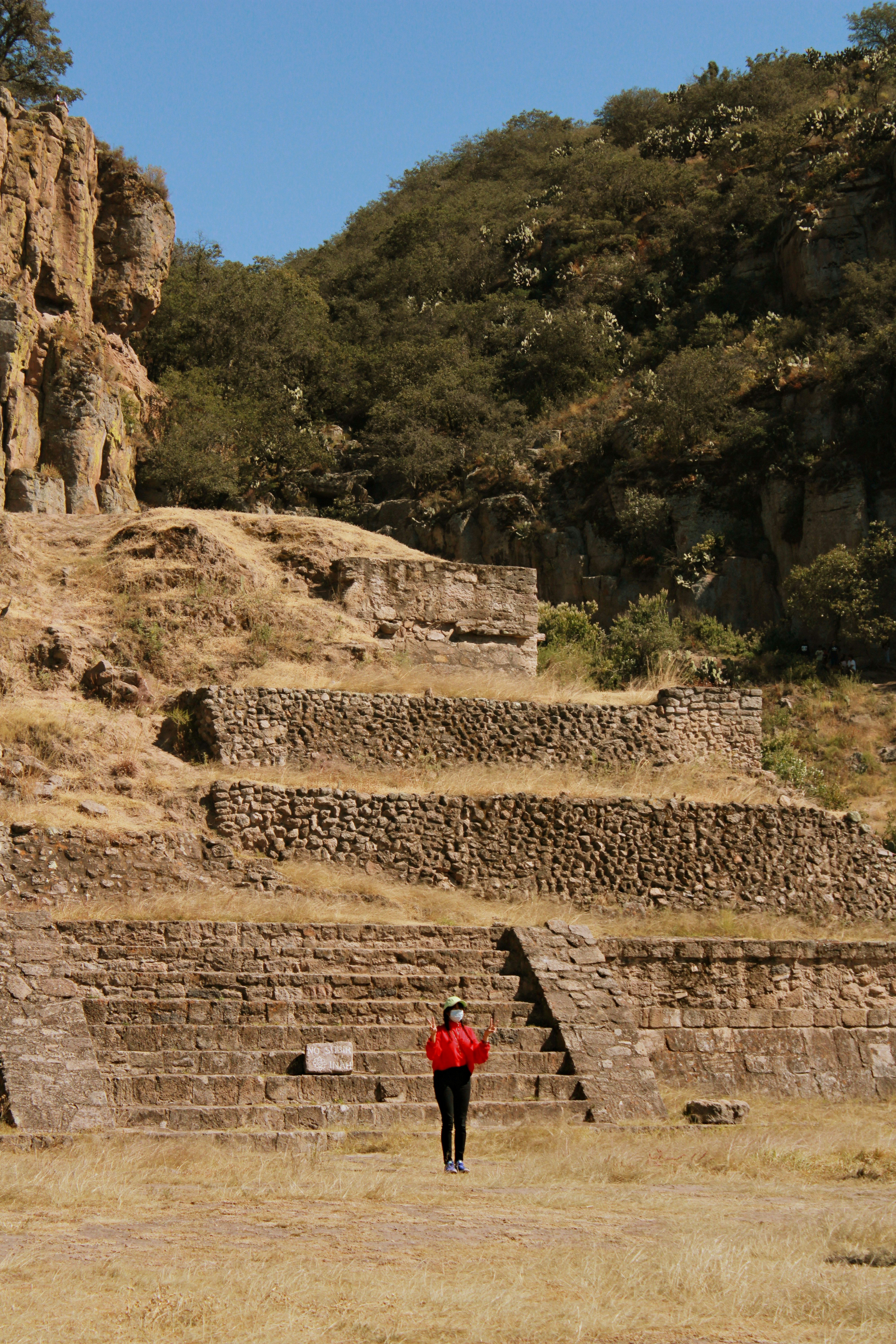 Person in a red jacket walking towards ancient stone terraces under a clear blue sky. The rugged landscape showcases the remnants of a historical site.