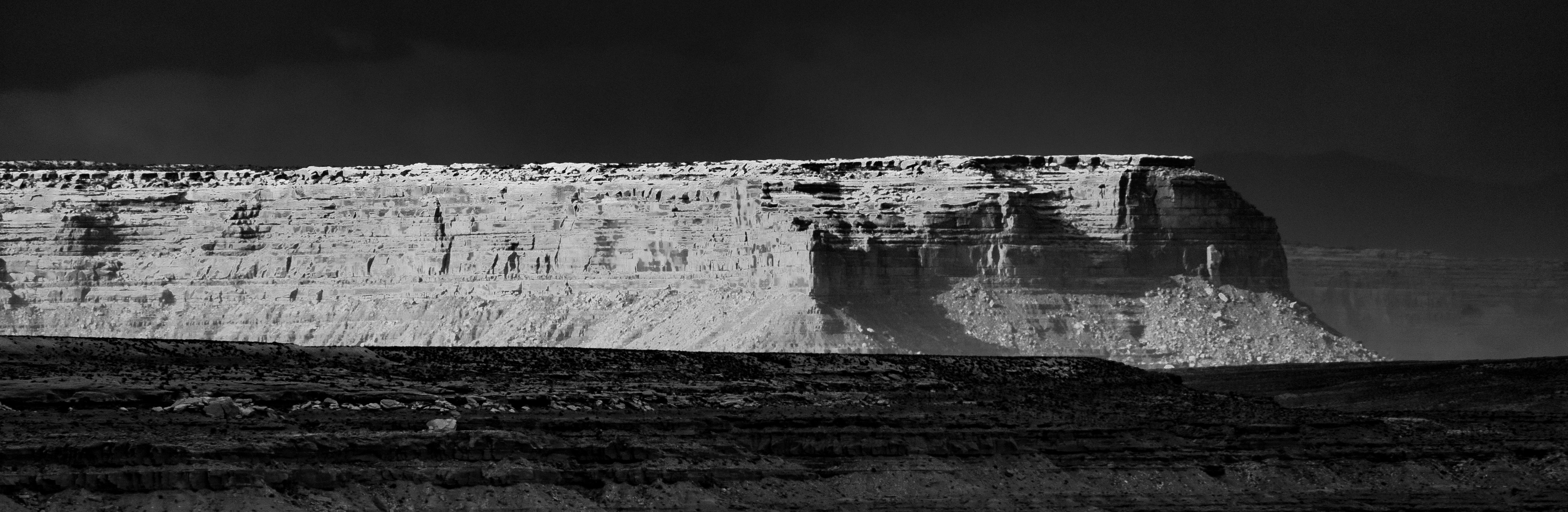 Monochrome landscape featuring a flat-topped mesa along the horizon against a barren foreground. The stark geometric forms and tonal contrast emphasize the scene's austere mood.