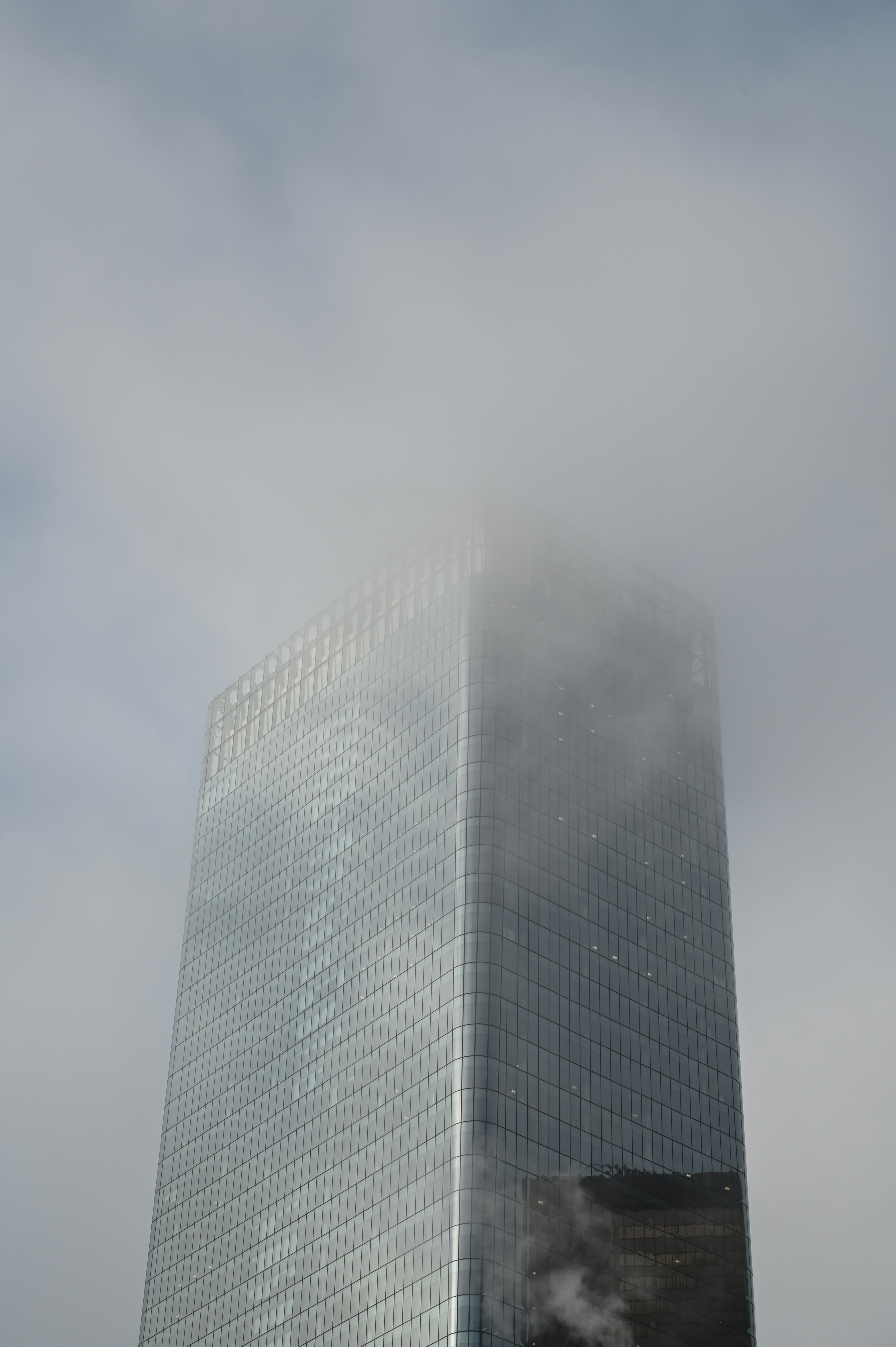 A modern skyscraper partially obscured by swirling clouds, creating a mysterious atmosphere. The reflective glass facade captures the soft light of the sky.